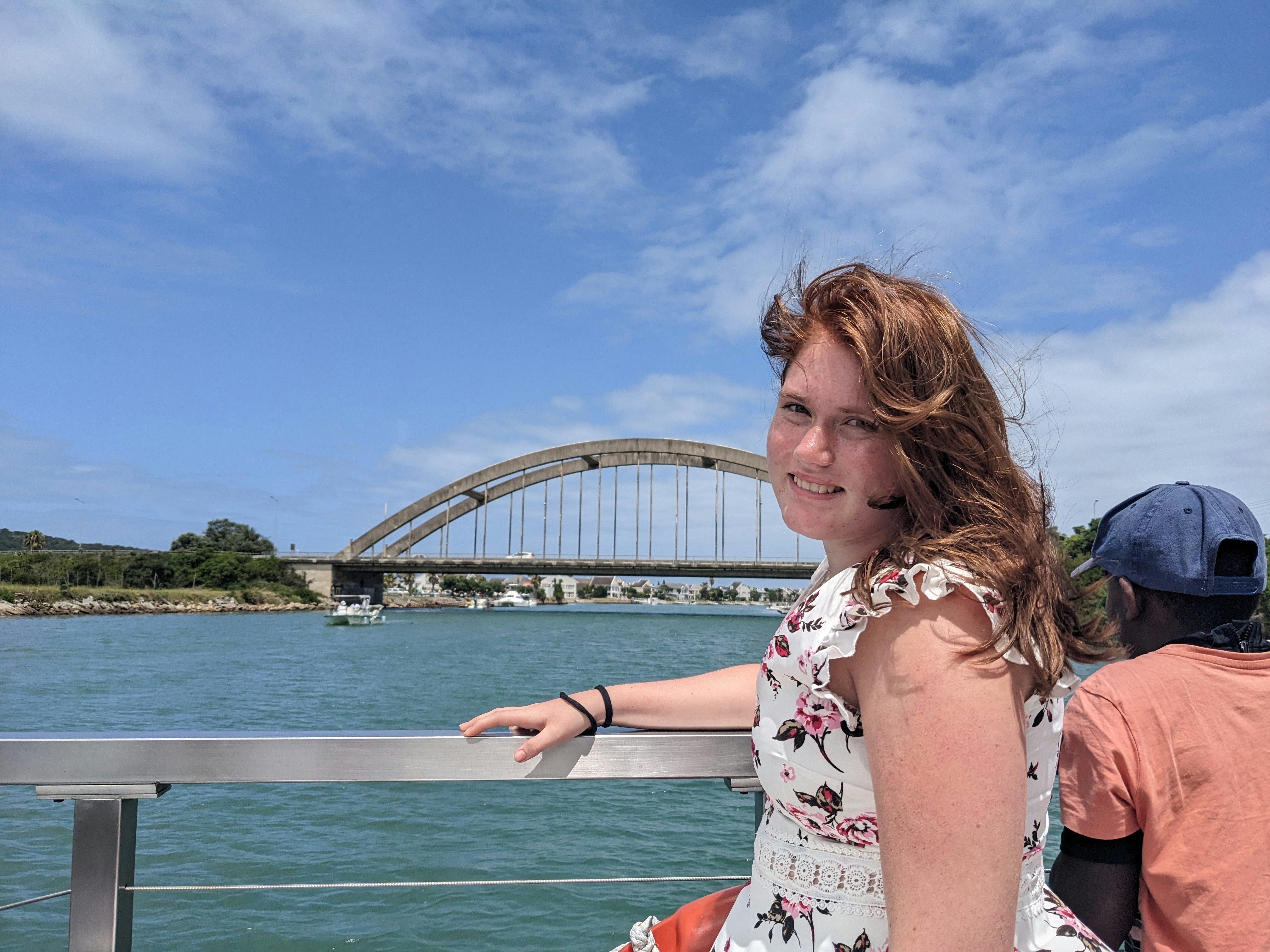 Woman in floral dress on a boat with an arch bridge in the background under a blue sky.