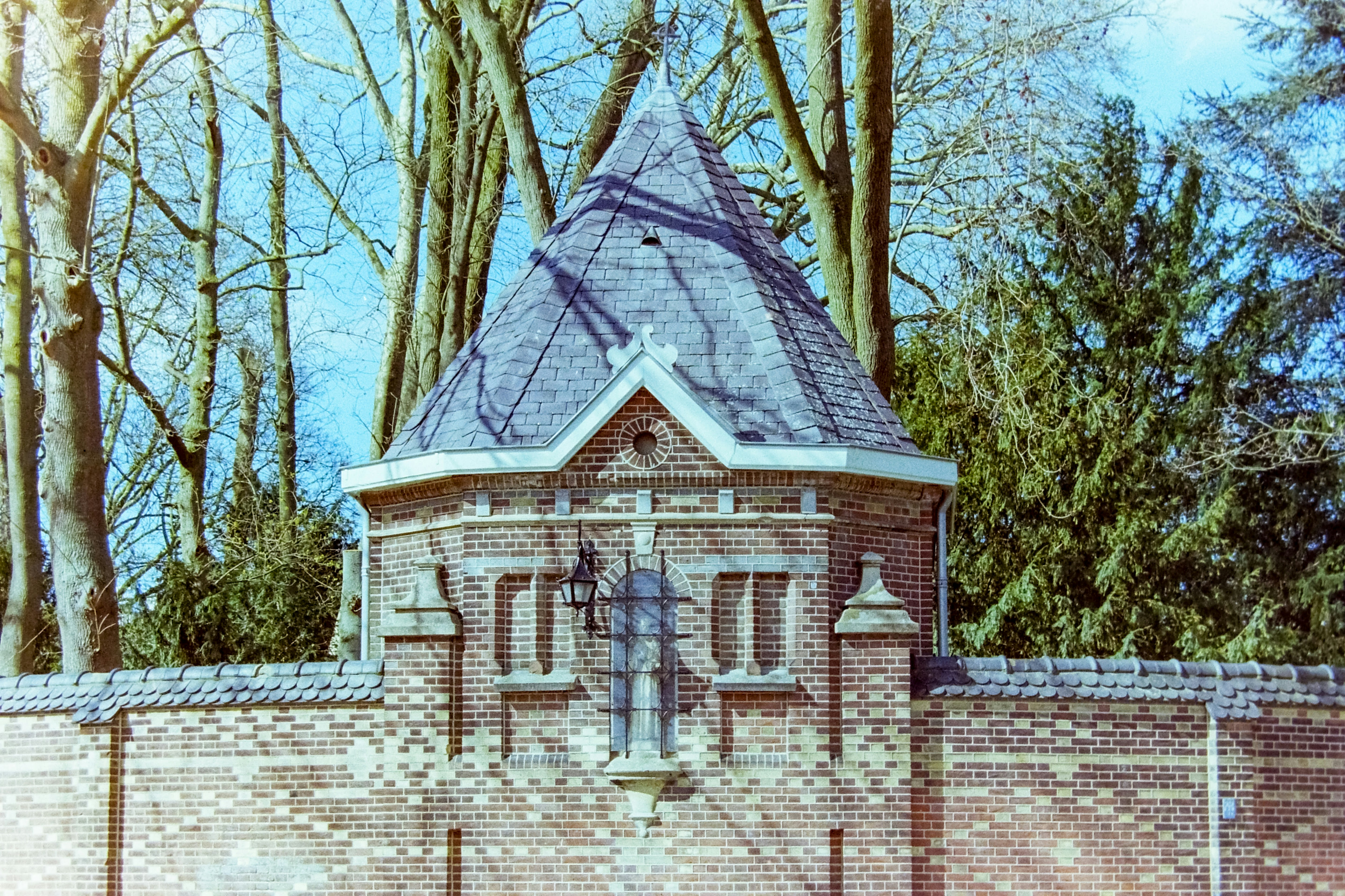 Statue mounted on a brick wall with a turreted roof, framed by tall leafless trees. The cool-toned scene evokes a quiet monument in a secluded courtyard.
