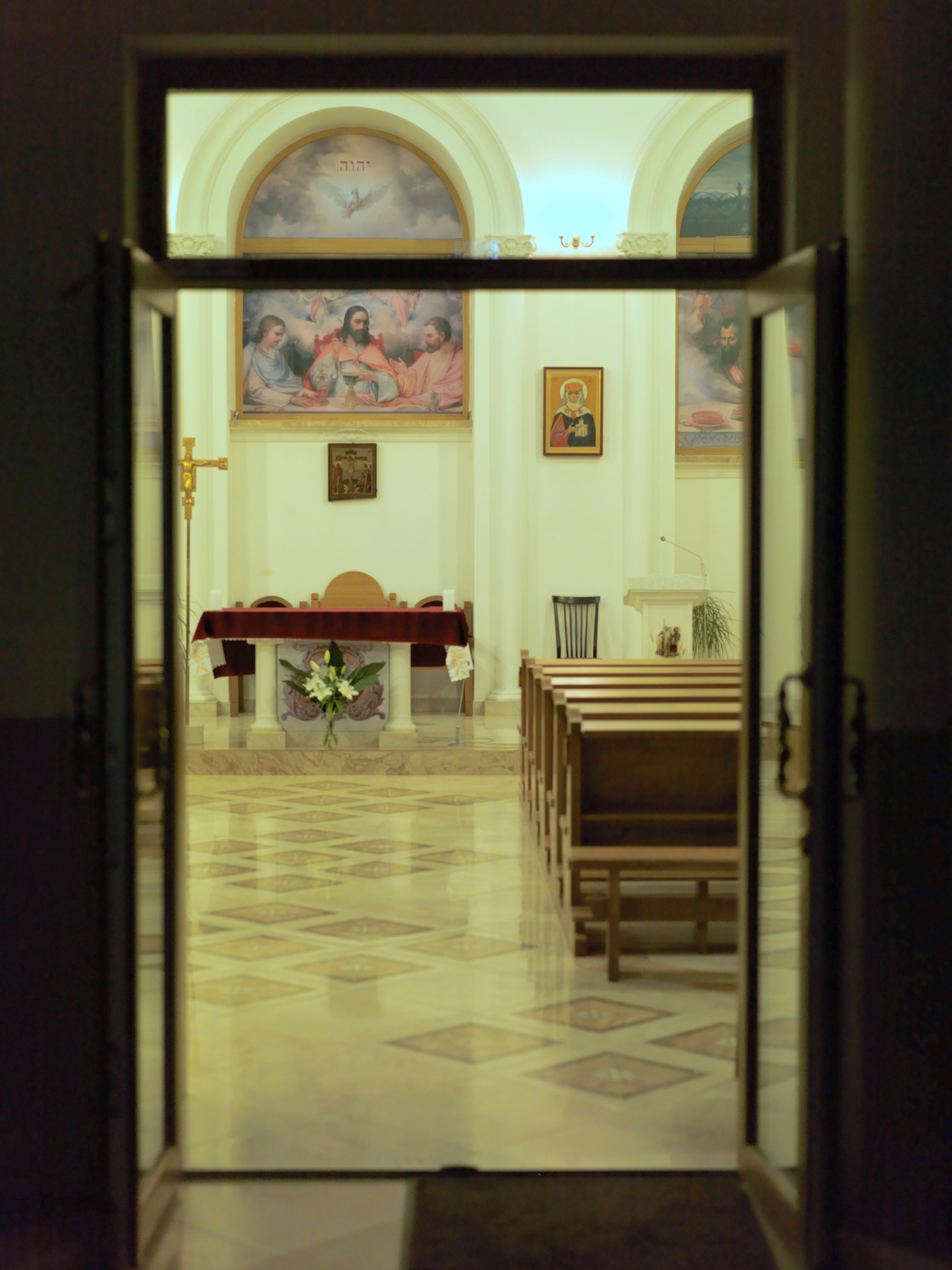View into a serene church interior featuring ornate paintings and a simple altar, framed by an open doorway.