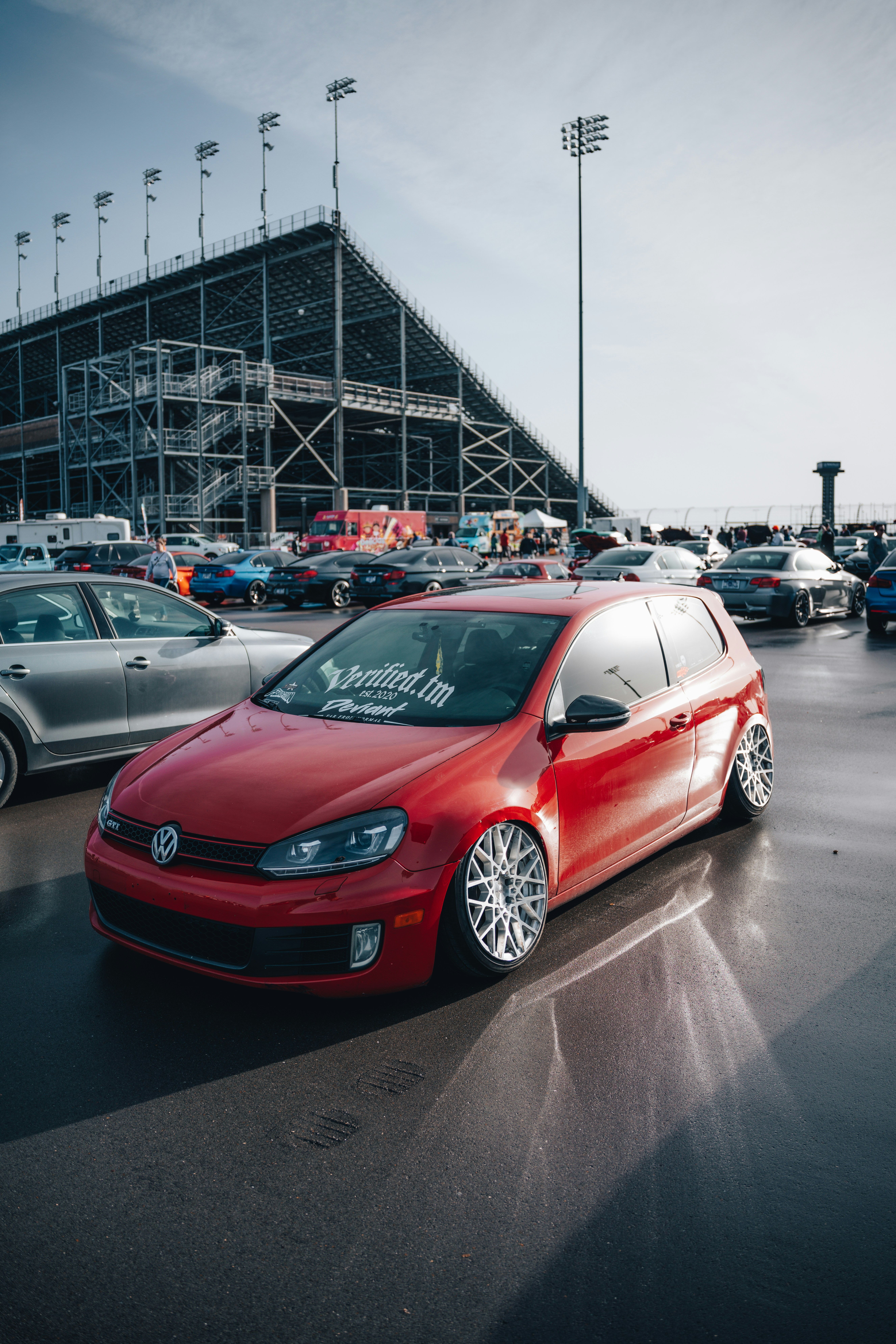 Red Volkswagen GTI at a sunlit car meet with a grandstand backdrop