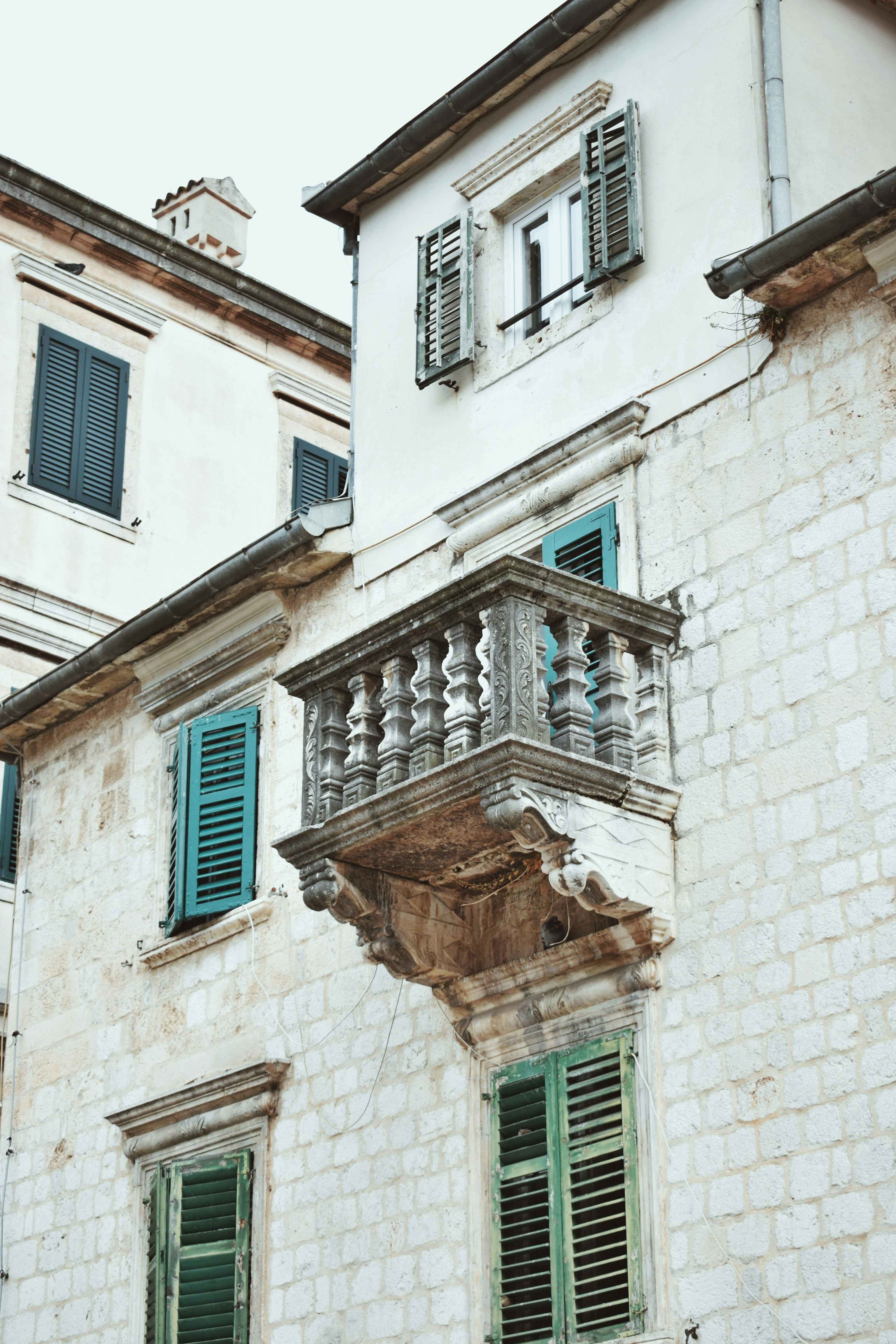 Architectural detail of a historic building featuring a decorative balcony and green shutters. The weathered stone walls tell a story of time gone by.