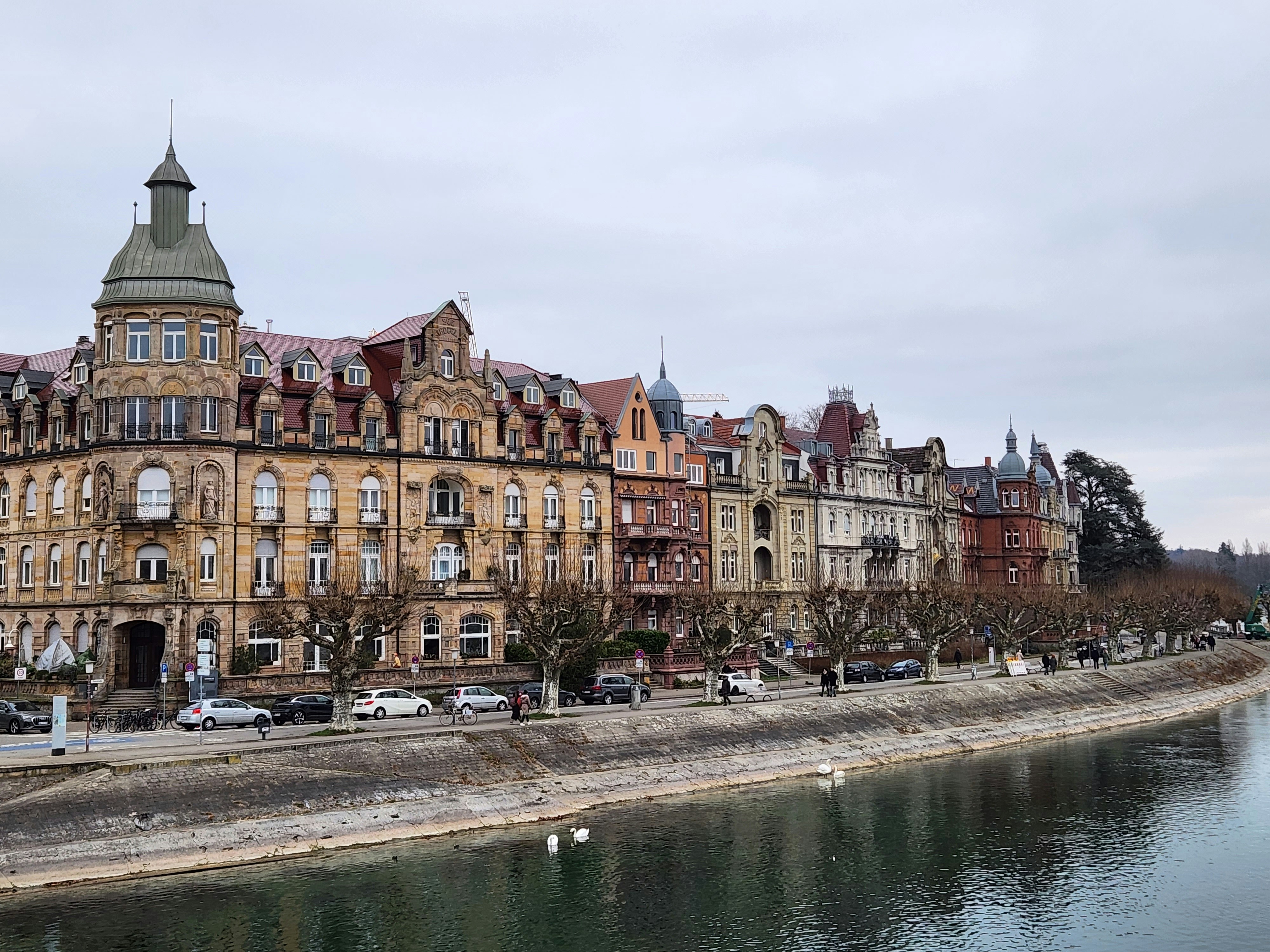 Historic European buildings line a riverbank under an overcast sky.