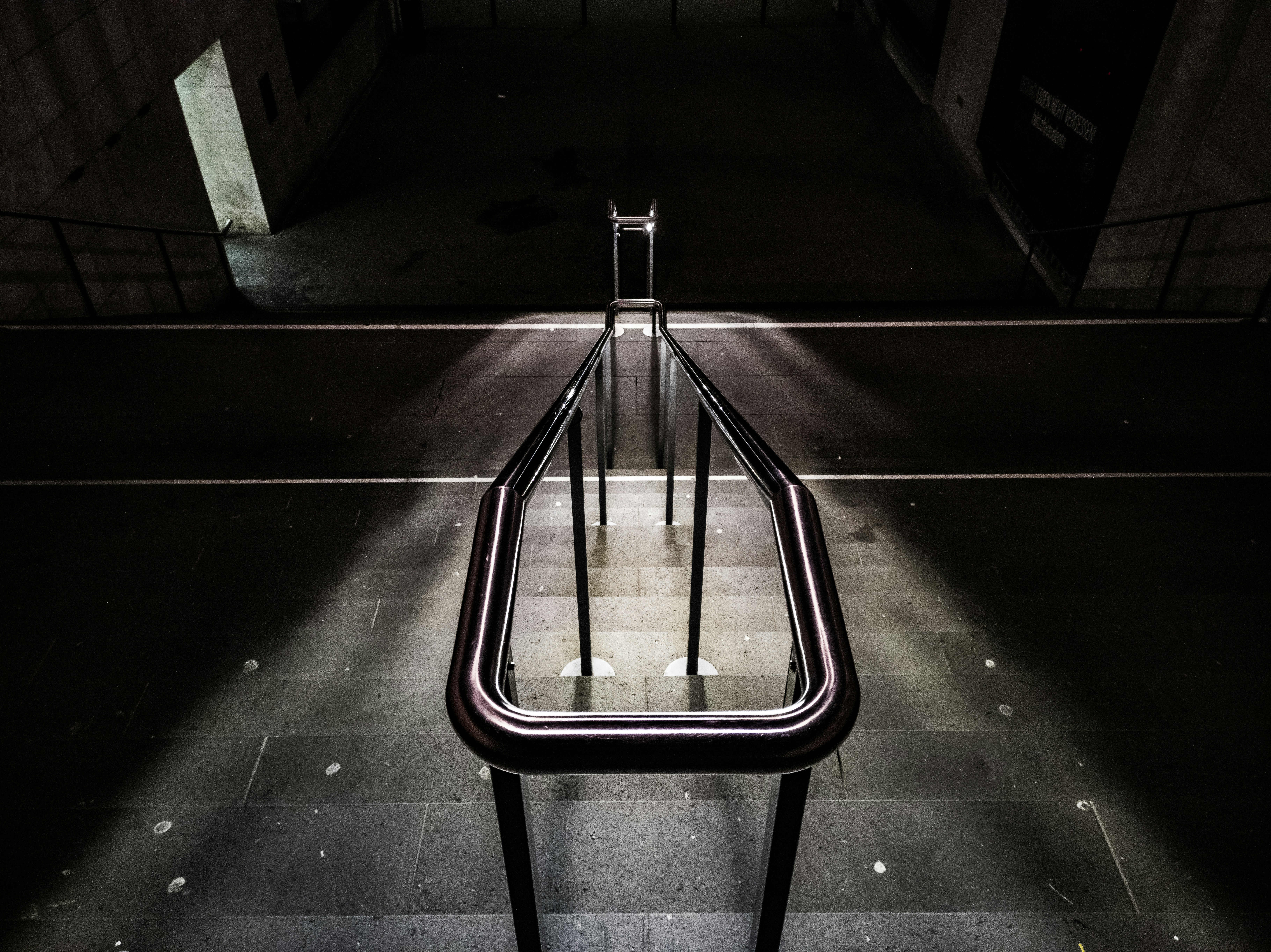 Dark urban stairwell with a central, lit handrail leading downward; concrete walls fade to shadow, highlighting the glossy guard rails and illuminated steps.