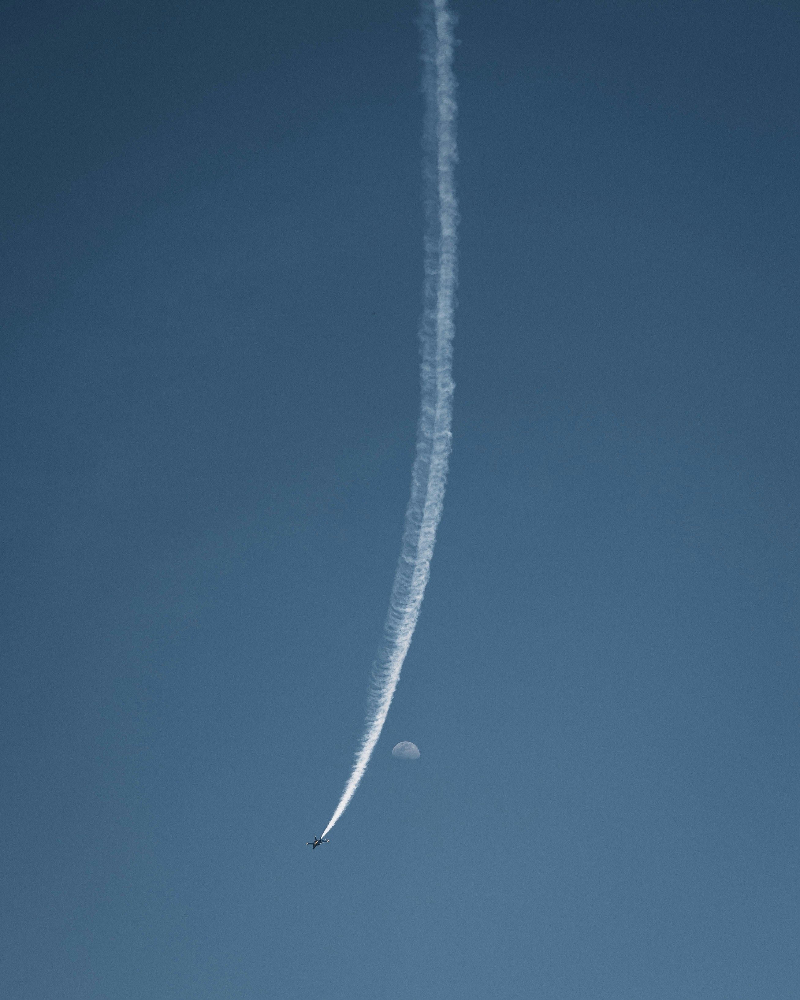 An airplane trails a wispy contrail against a clear blue sky, with the crescent moon subtly appearing in the background.