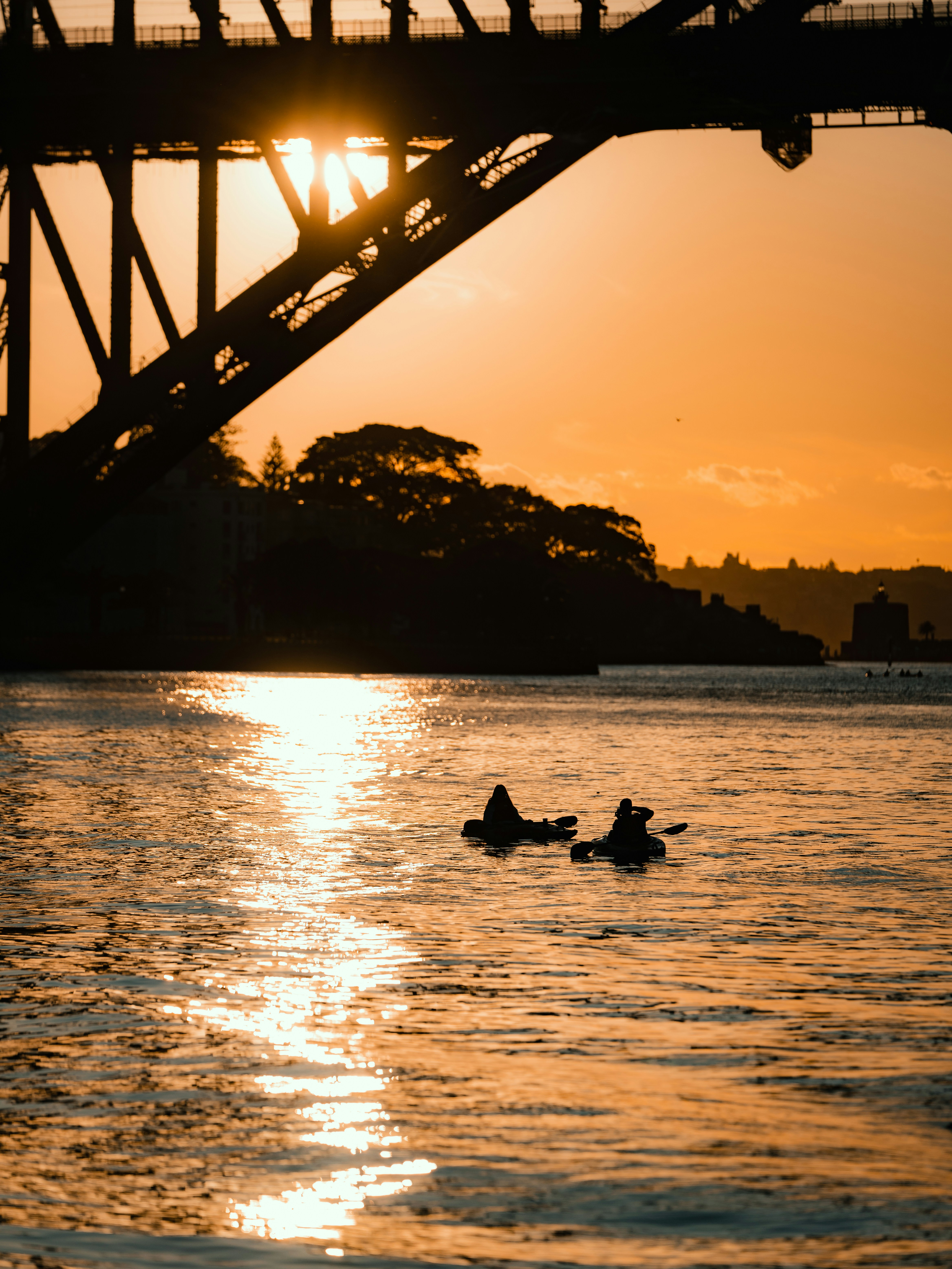 Two kayakers gliding across a shimmering river as the sun sets behind a towering bridge, casting warm golden hues on the water.