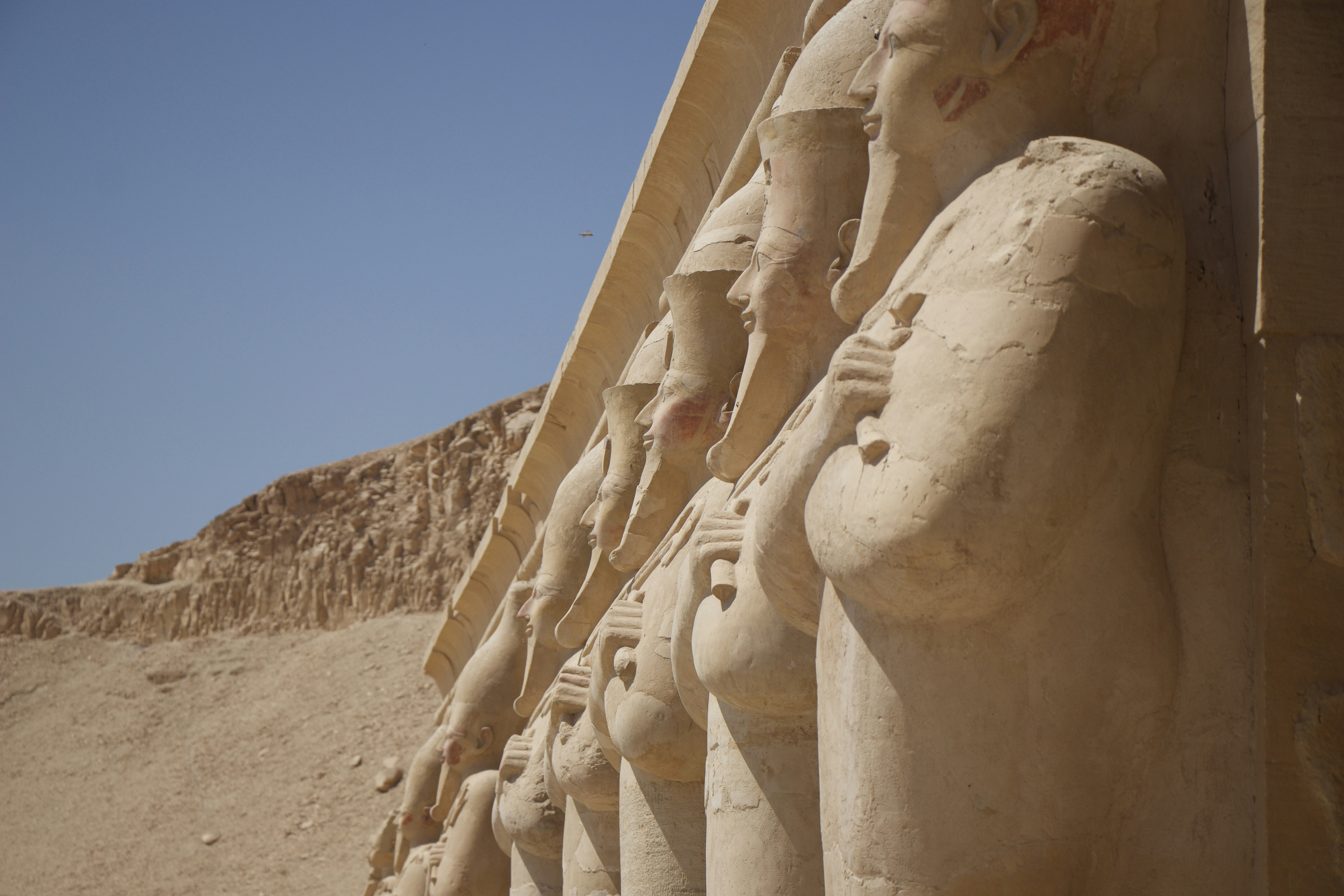 Ancient statues of pharaohs stand in a row against a desert backdrop under a clear blue sky.