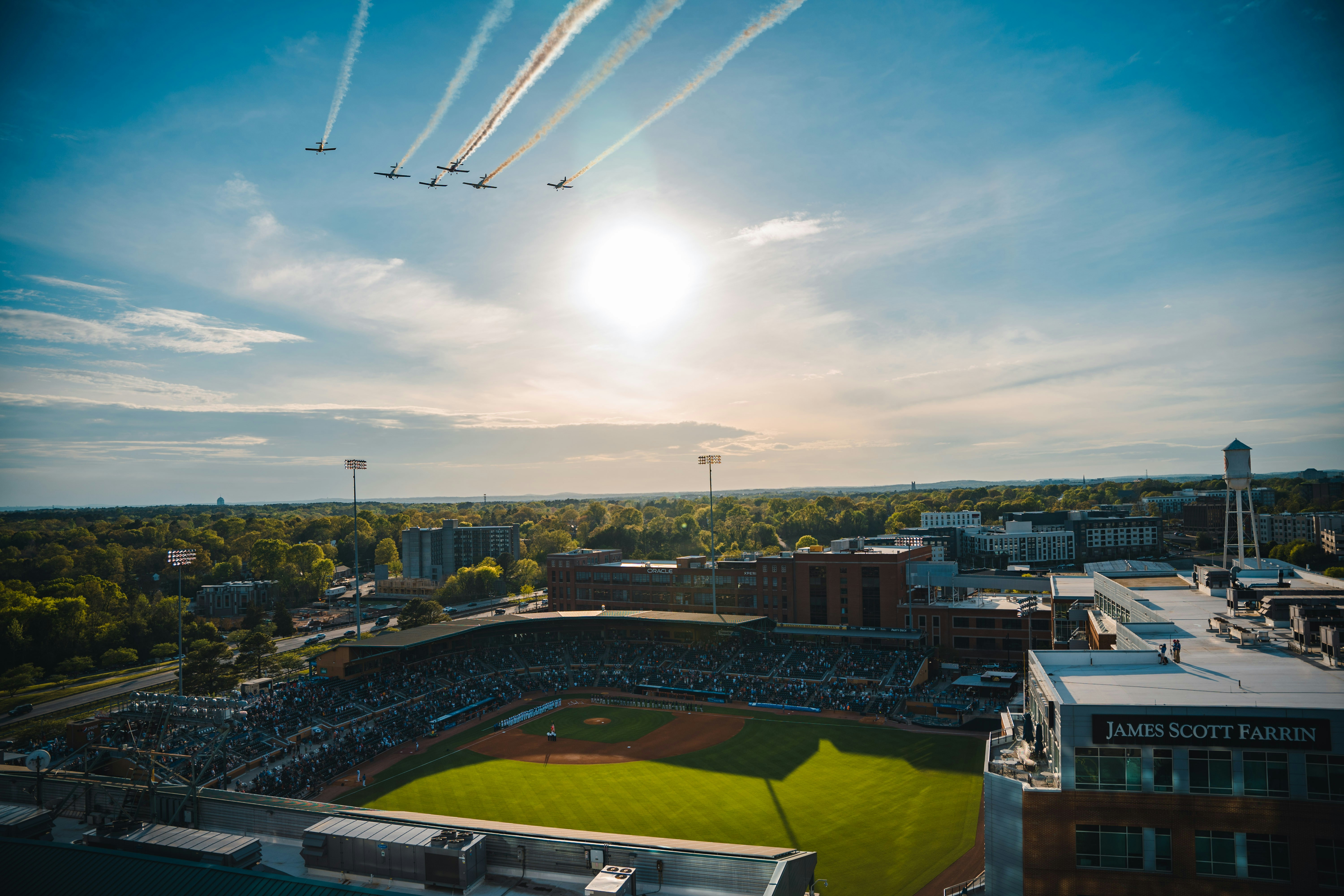 Jets streak across the sky over a sunlit baseball stadium with a vibrant field.