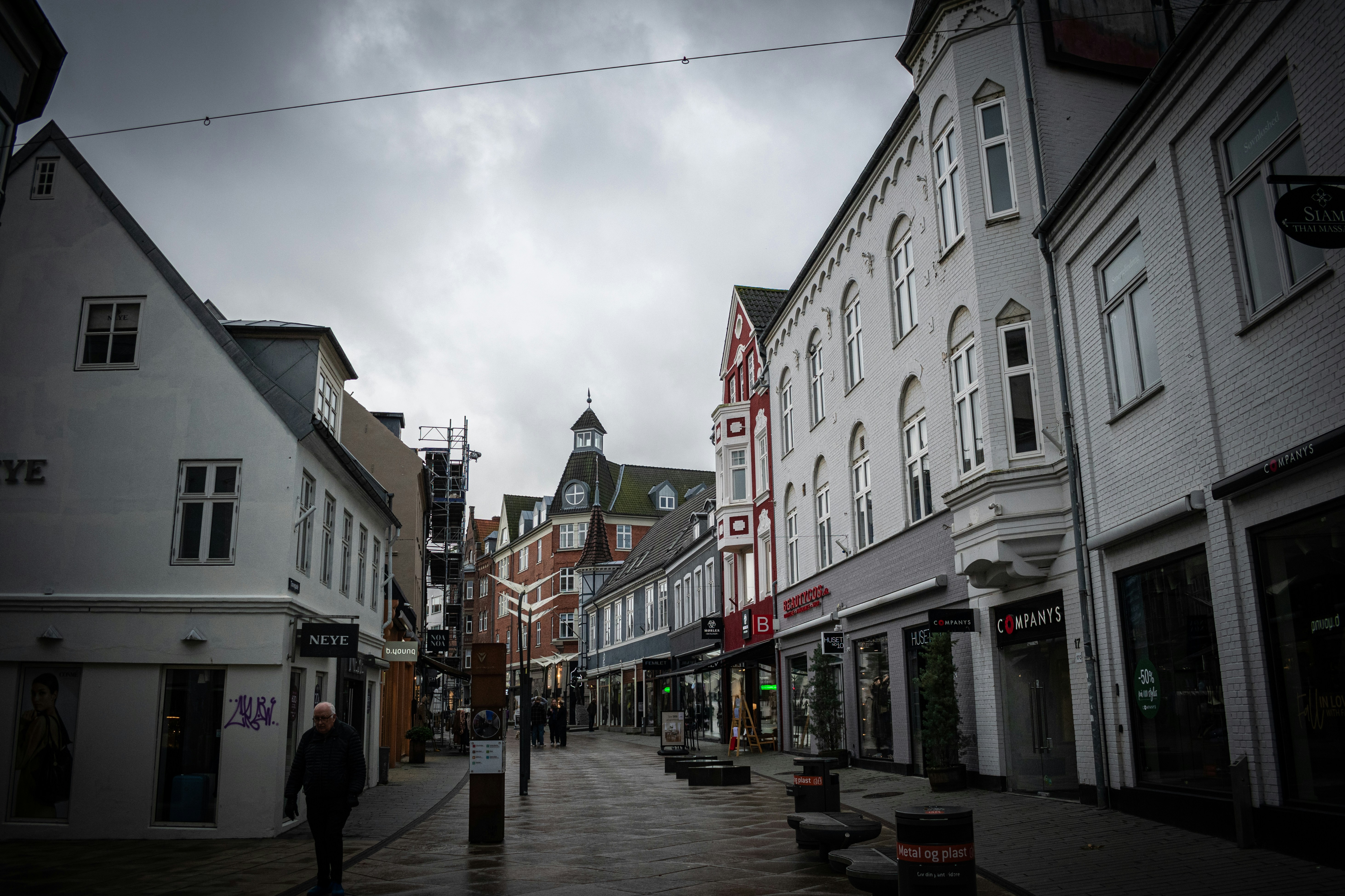 A quiet street scene showcasing charming architecture with a mix of modern and traditional styles, under a moody sky. People stroll leisurely, highlighting the urban atmosphere.