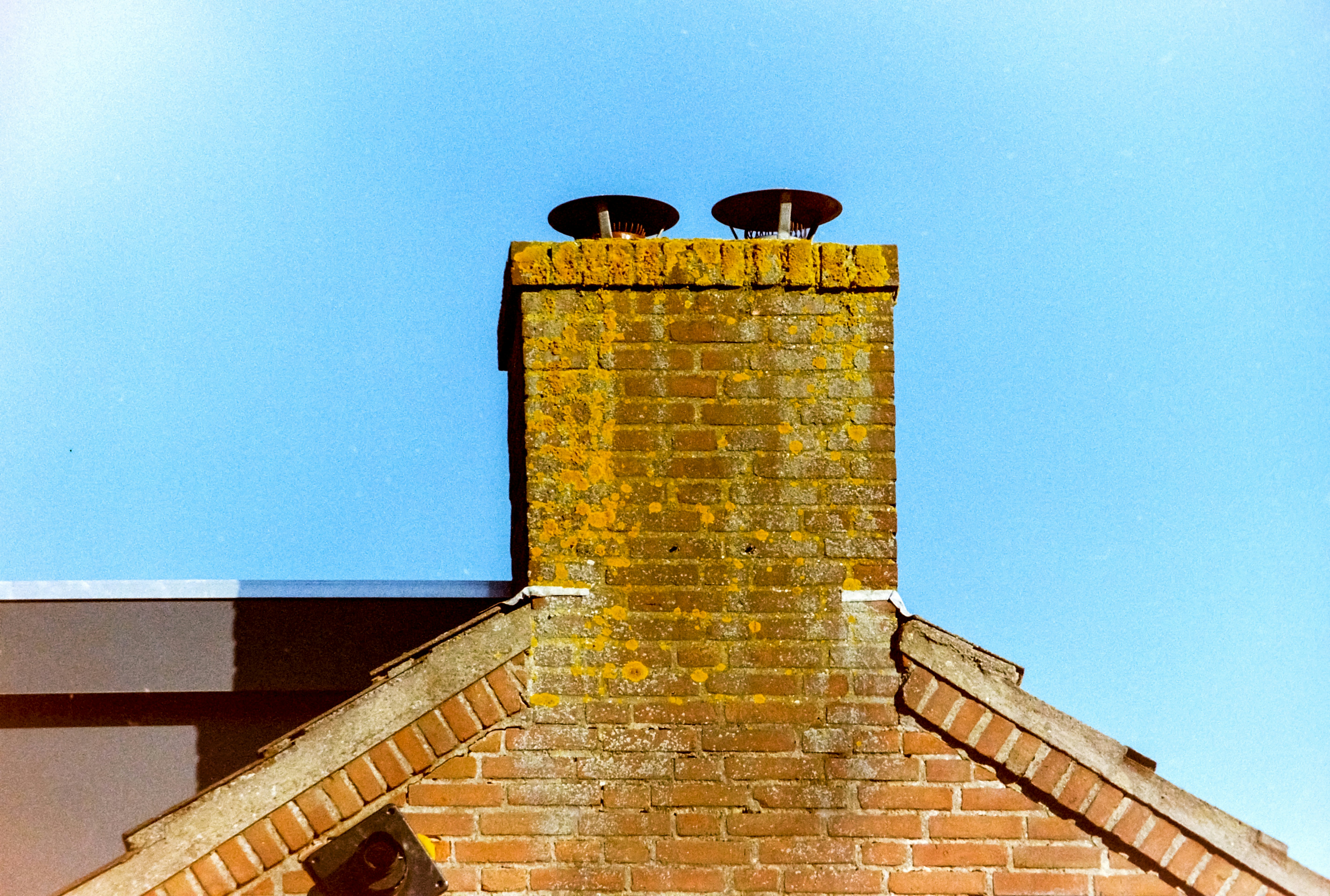 Weathered brick chimney adorned with moss, topped with two circular caps, set against a bright blue sky.
