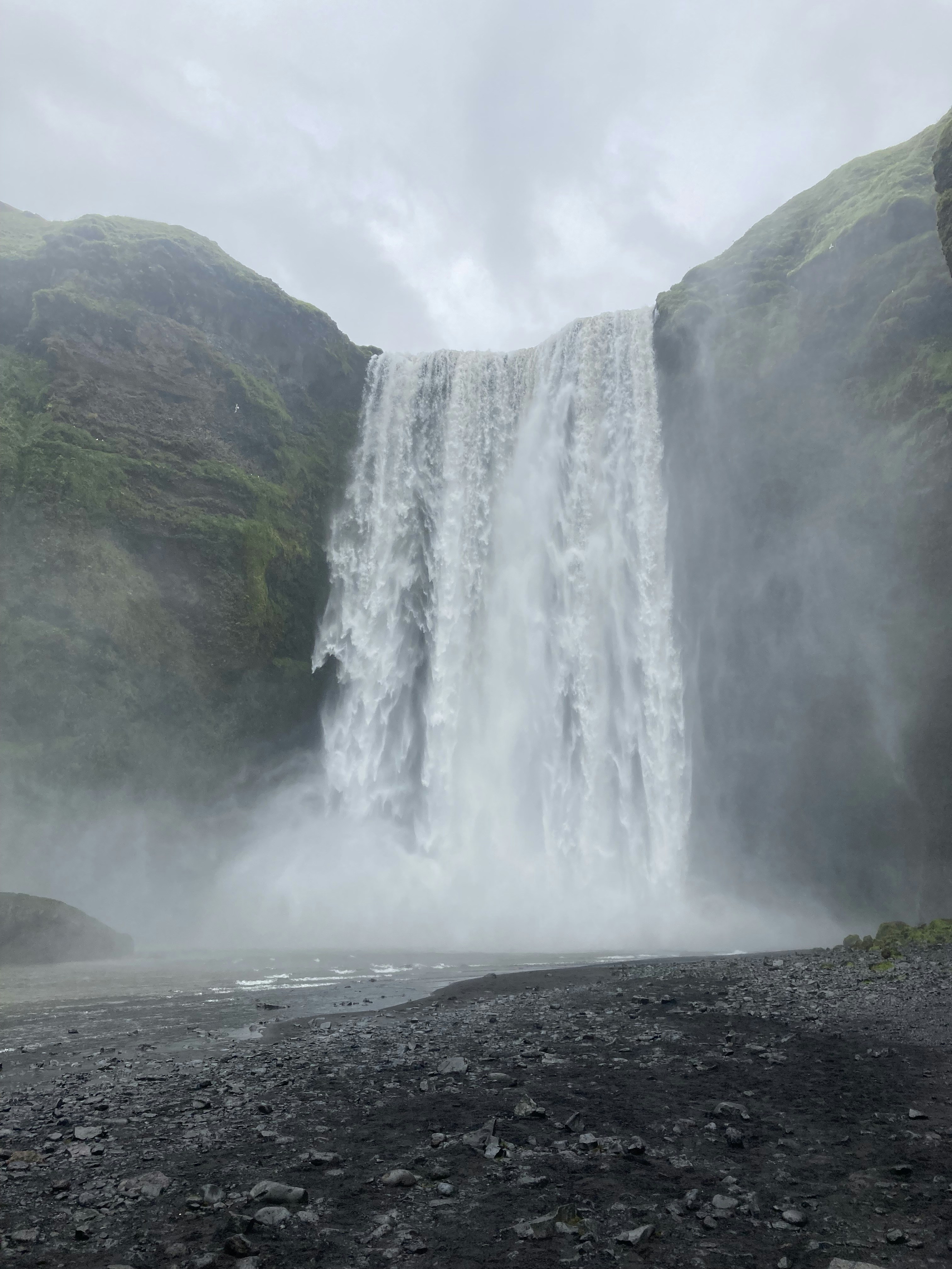 Waterfall curtain cascades between moss-covered cliffs, sending a veil of spray across a rocky riverbed.