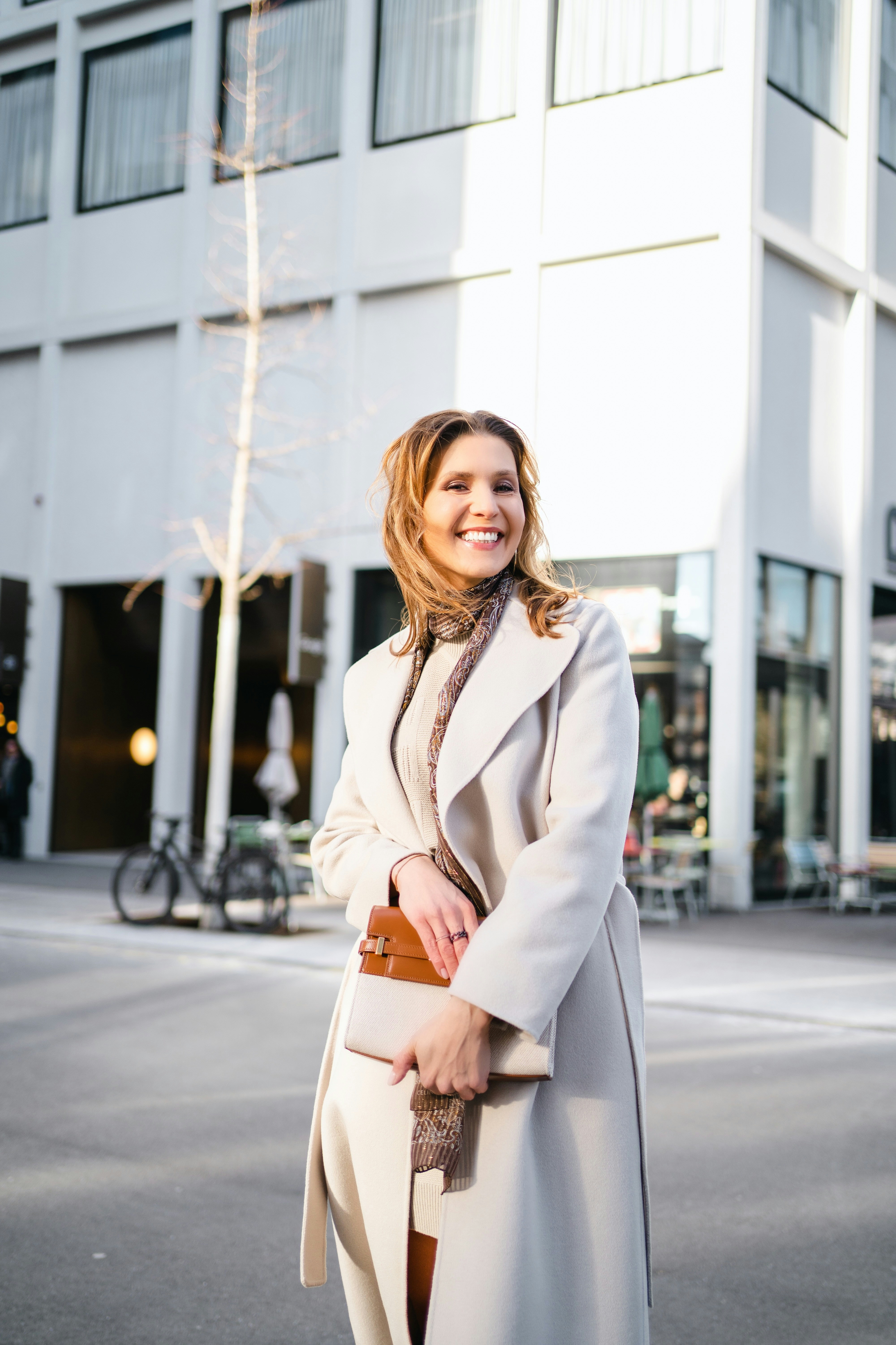 Smiling woman in a stylish coat stands against a modern urban backdrop, exuding confidence and warmth. The scene captures a blend of contemporary architecture and winter fashion.