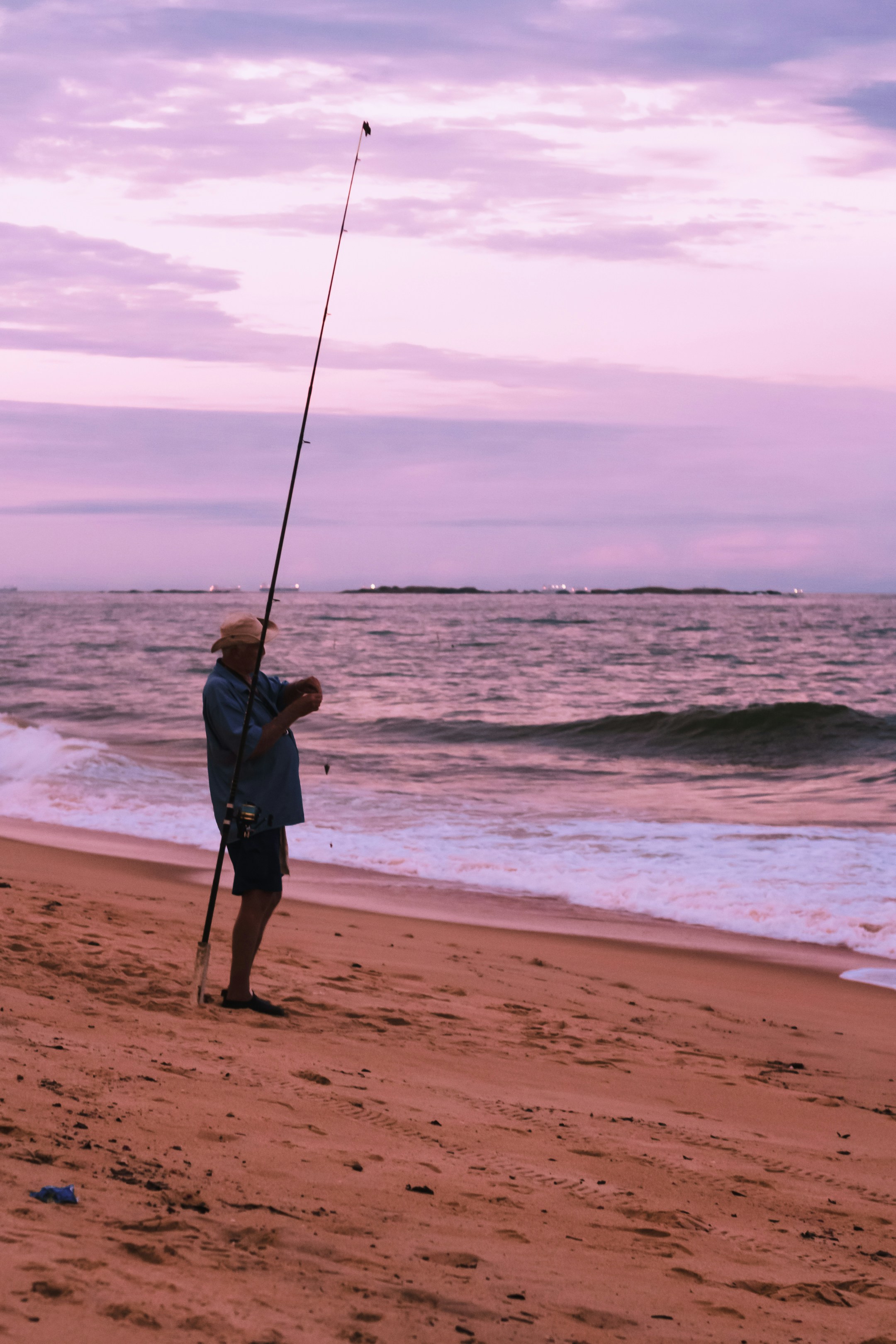 An elderly man stands on the shore, casting his fishing line into the tranquil ocean under a pastel sky. Waves gently lap at the sandy beach.