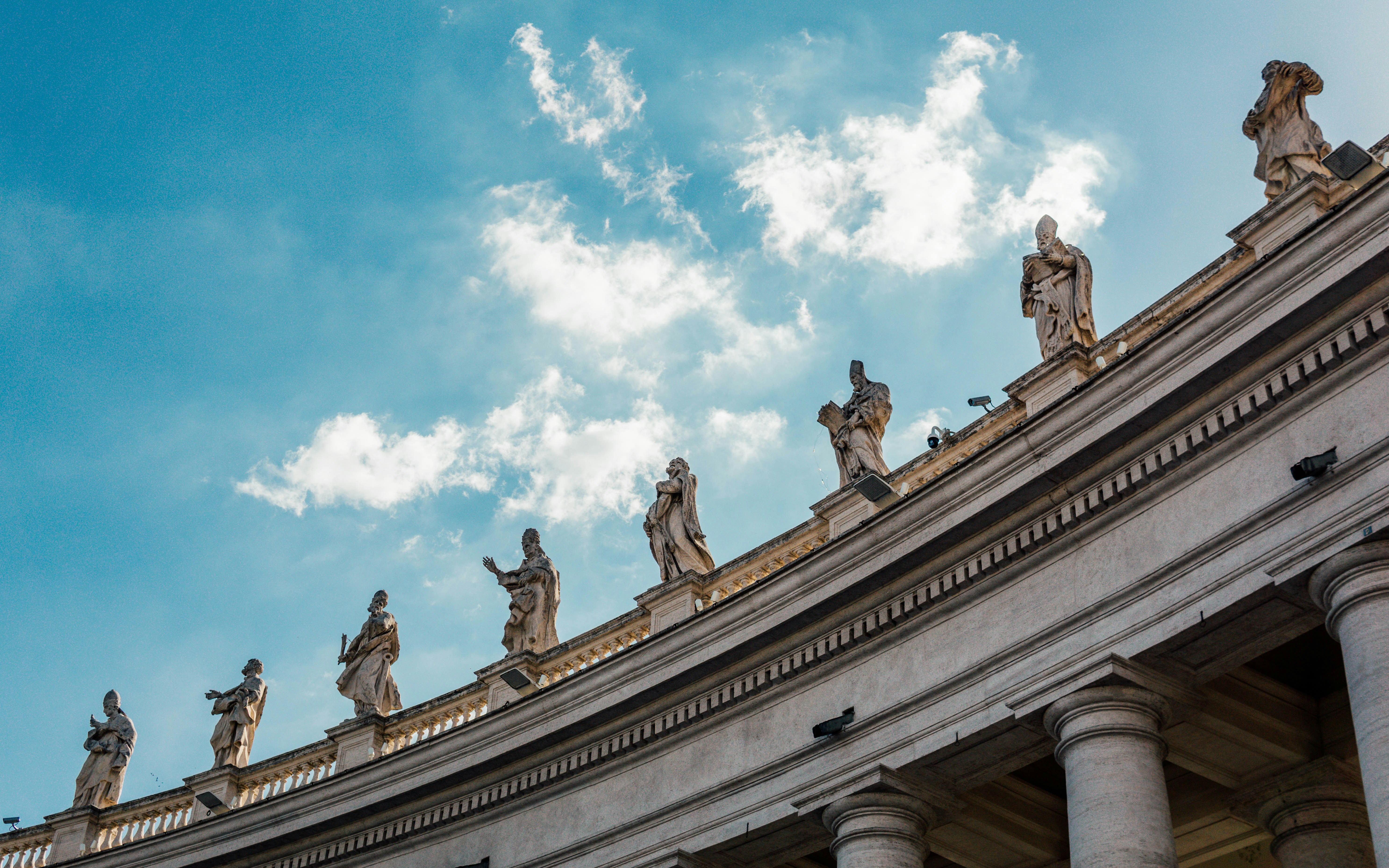 Row of statues atop a curved building facade against a bright blue sky with clouds.