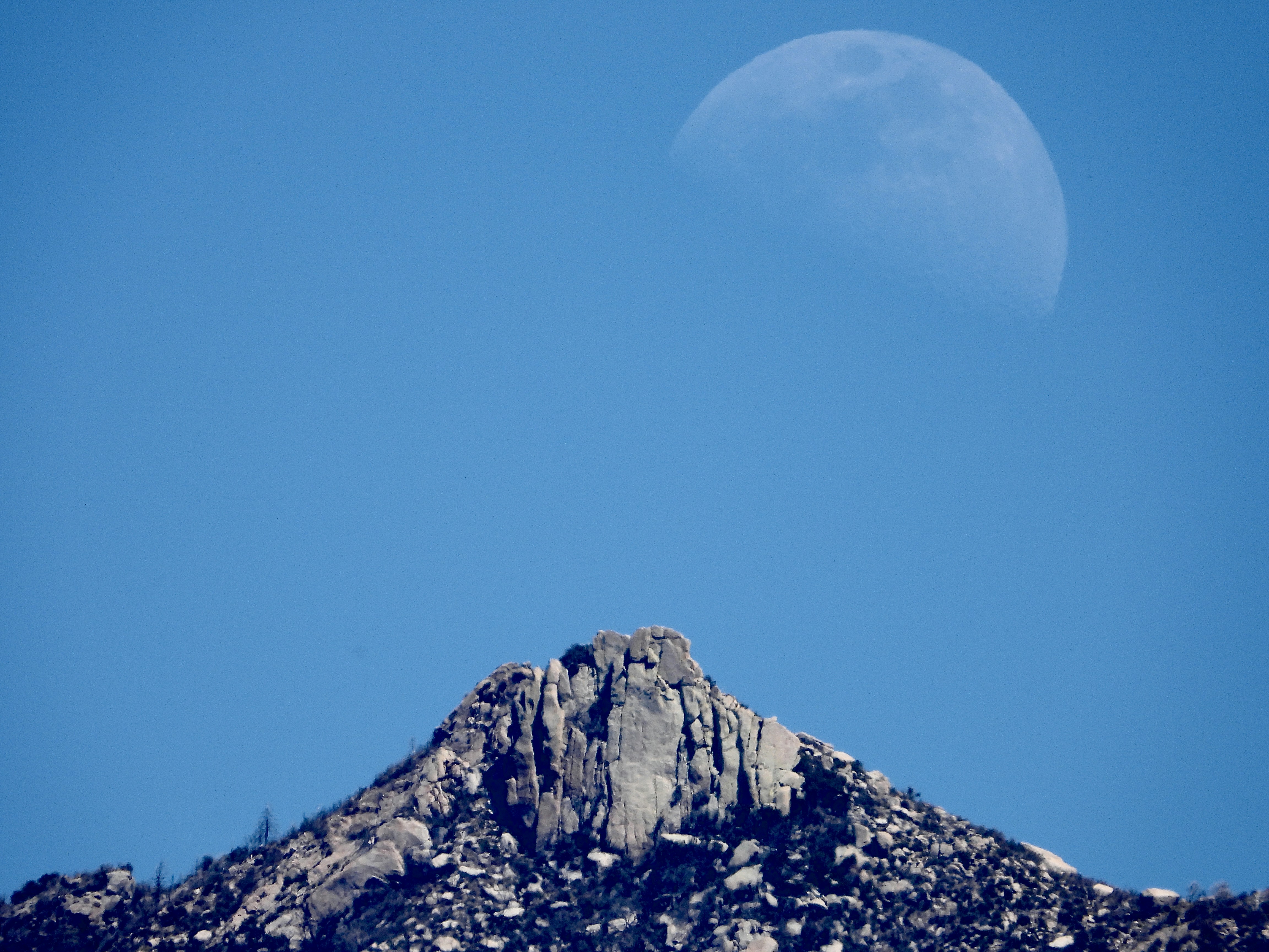 A rugged mountain peak stands resolute beneath a pale moon, creating a serene contrast against the clear blue sky.