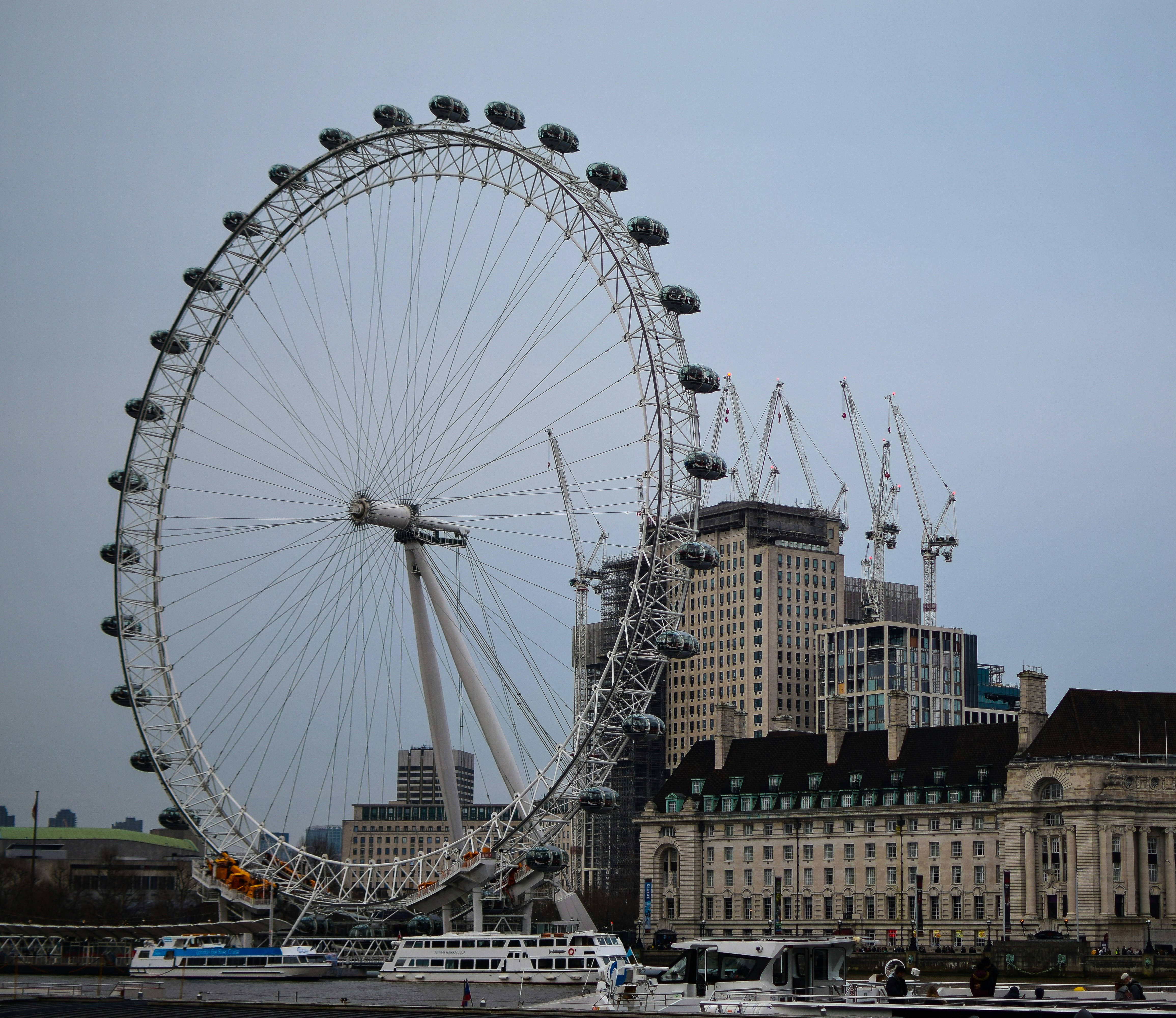 London Eye with towering cranes and historic architecture on a cloudy day.