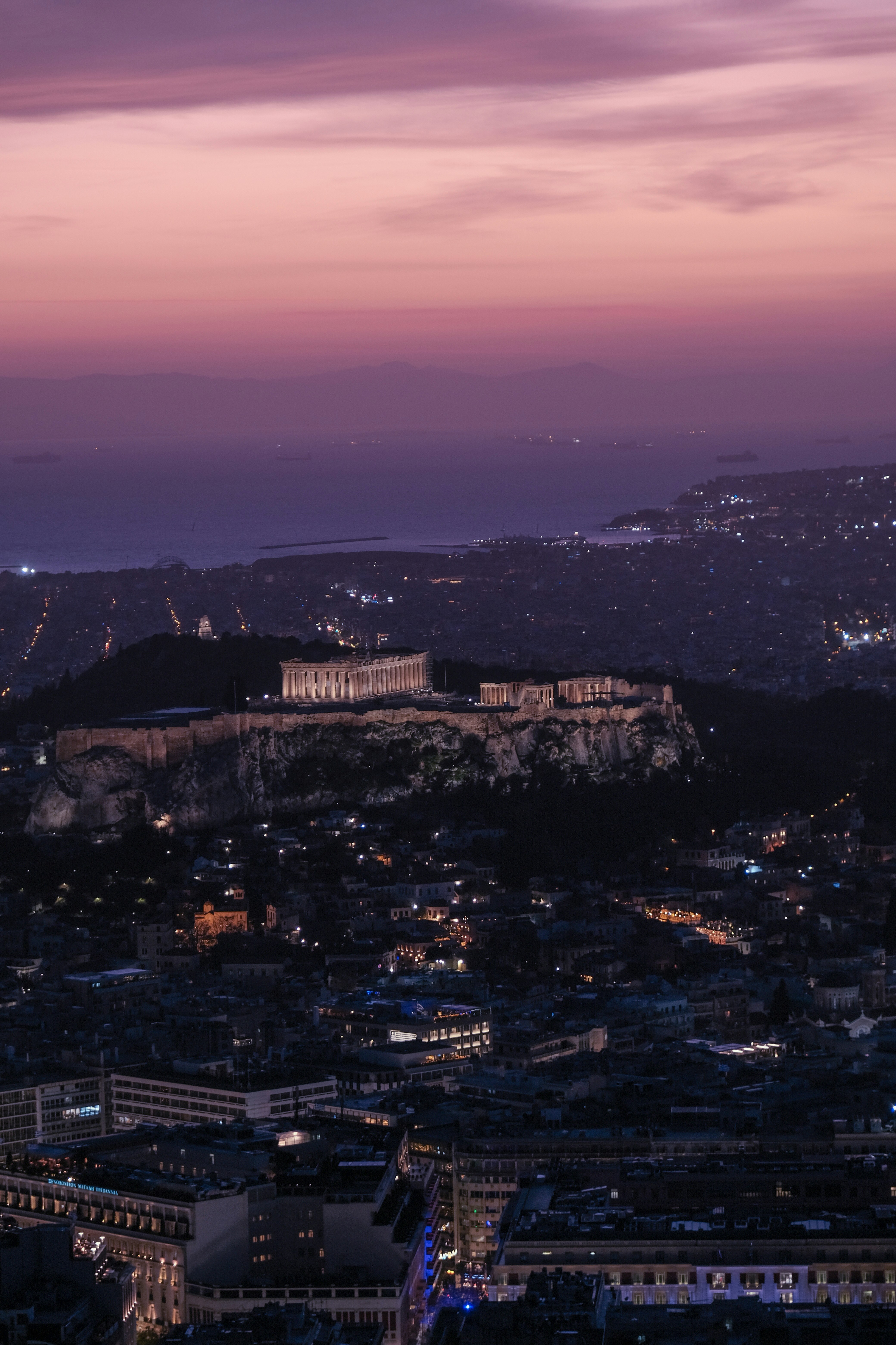 The Acropolis stands majestically against the twilight sky, surrounded by the twinkling lights of Athens below. A serene view capturing the essence of history and modernity.