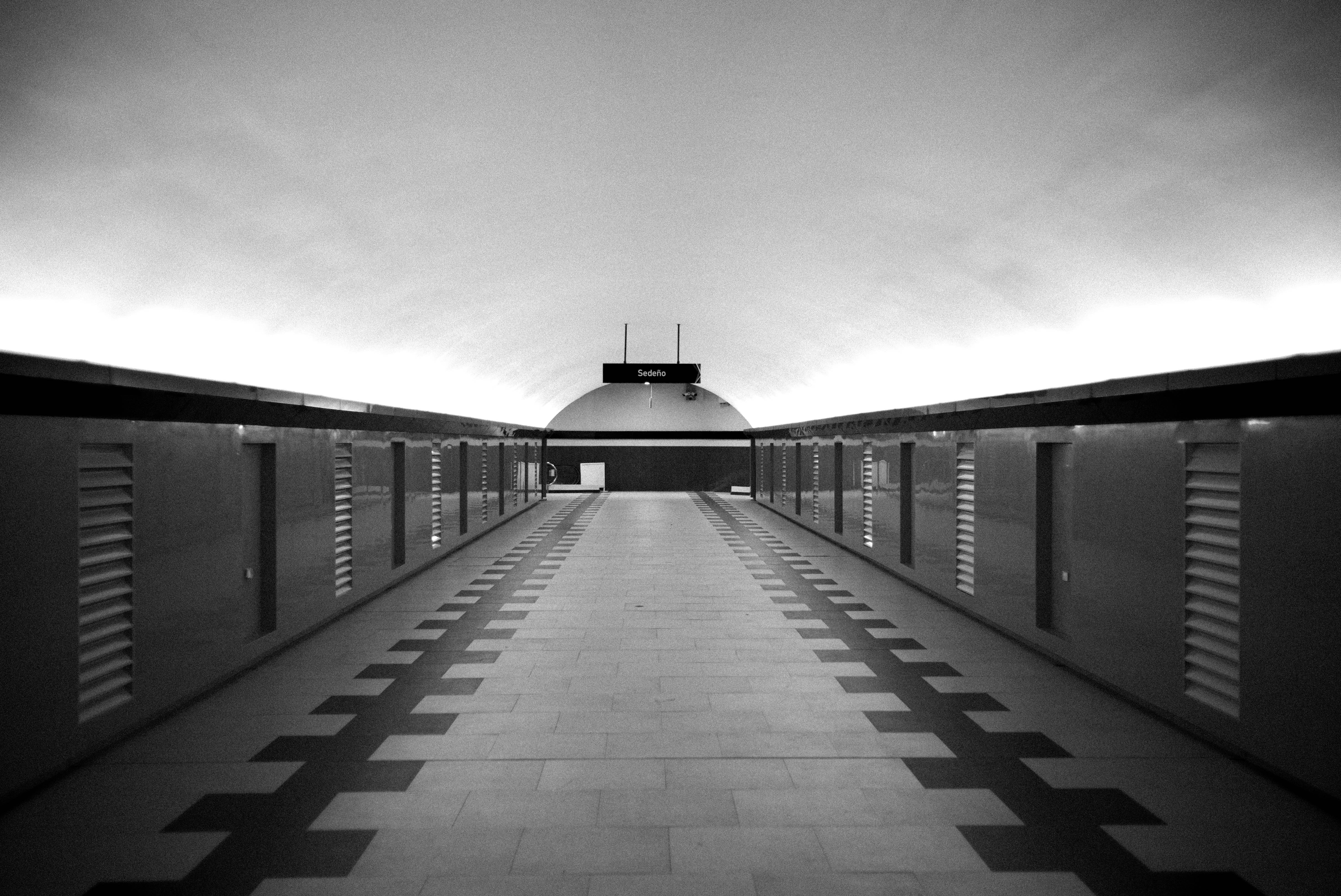 Monochrome hallway with symmetrical lines leading to a domed structure under a cloudy sky.