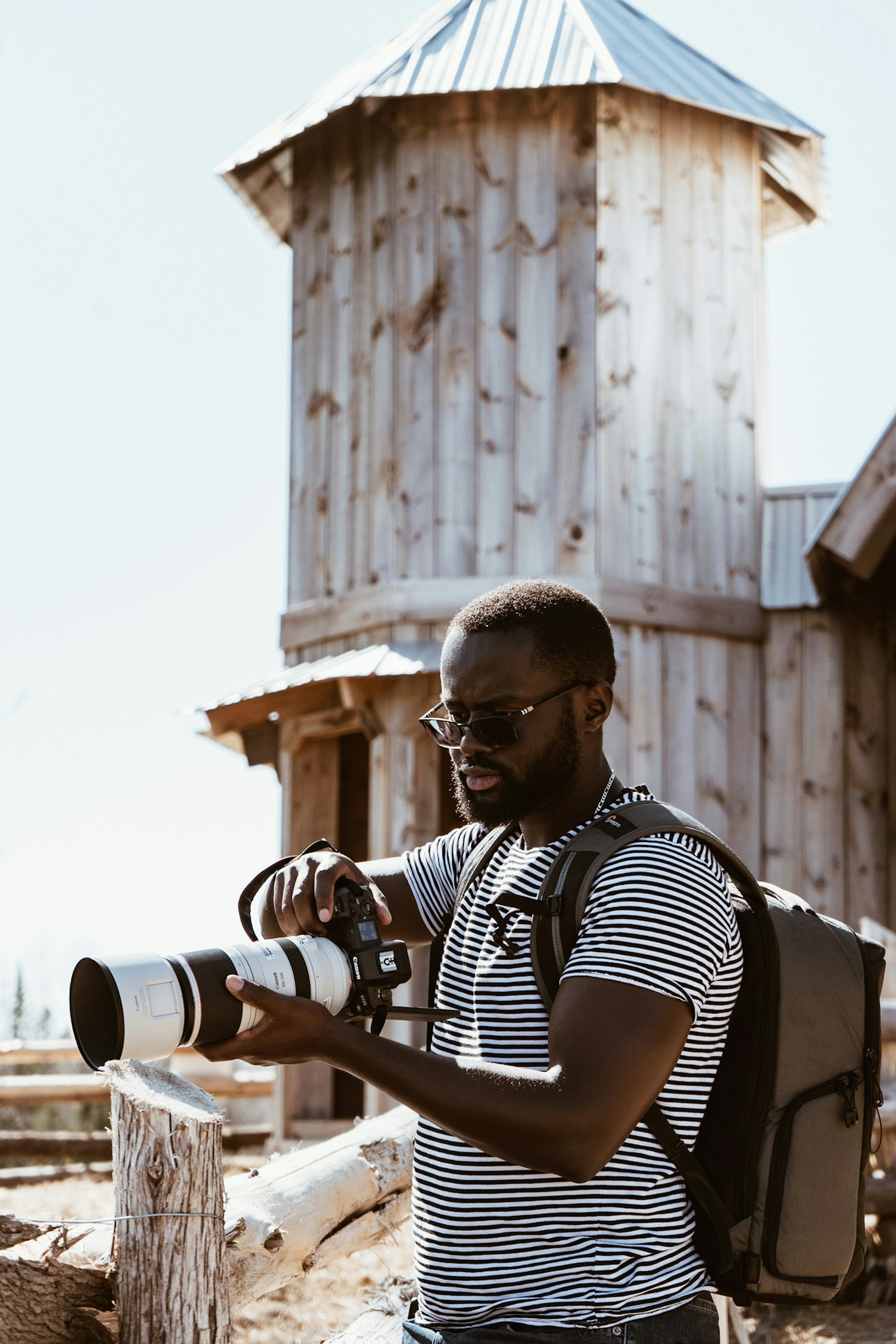 Photographer adjusting camera settings near a wooden structure on a sunny day.