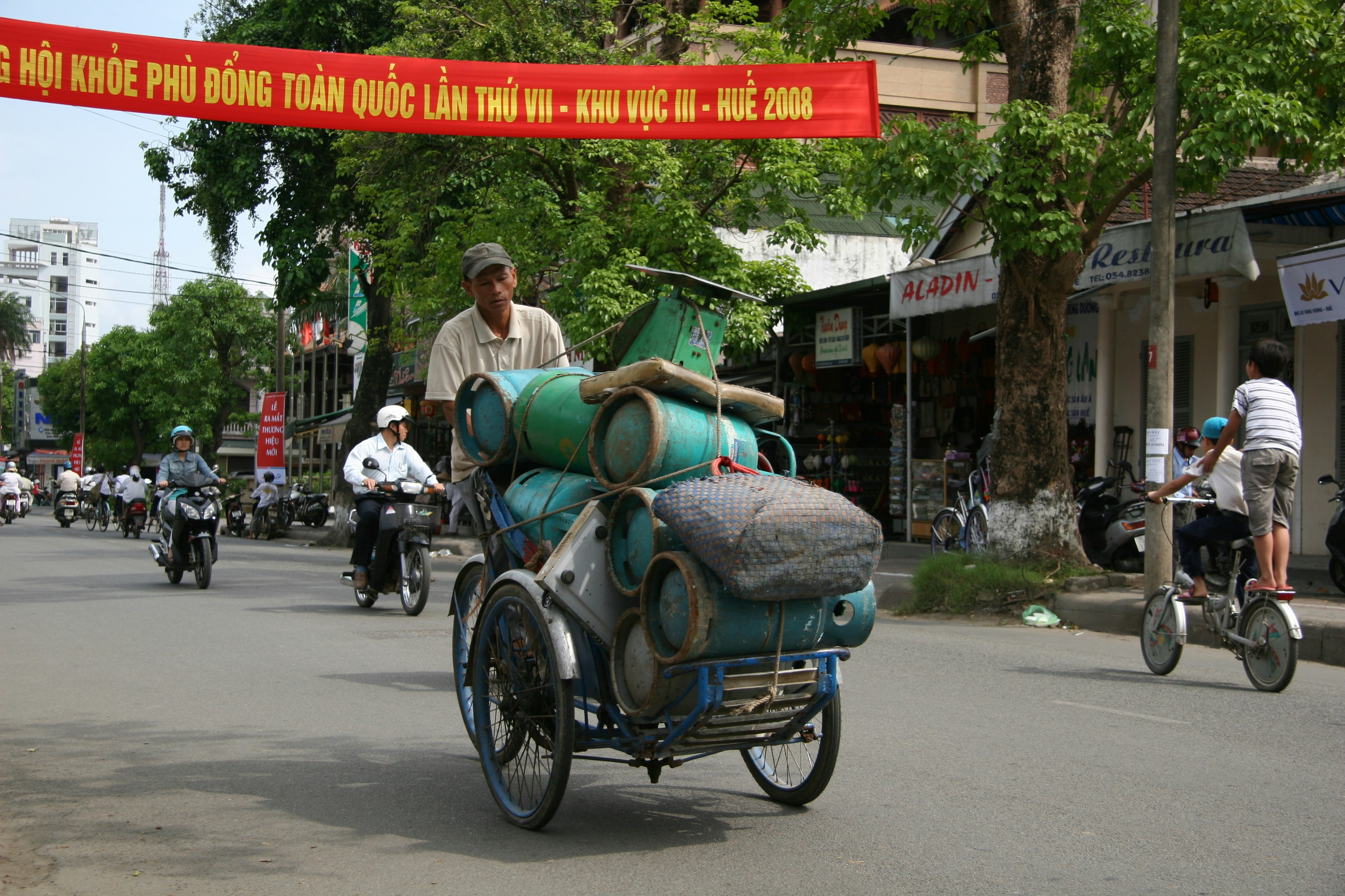 Man pushing a heavily loaded tricycle cart along a busy street under a red banner in a bustling city.