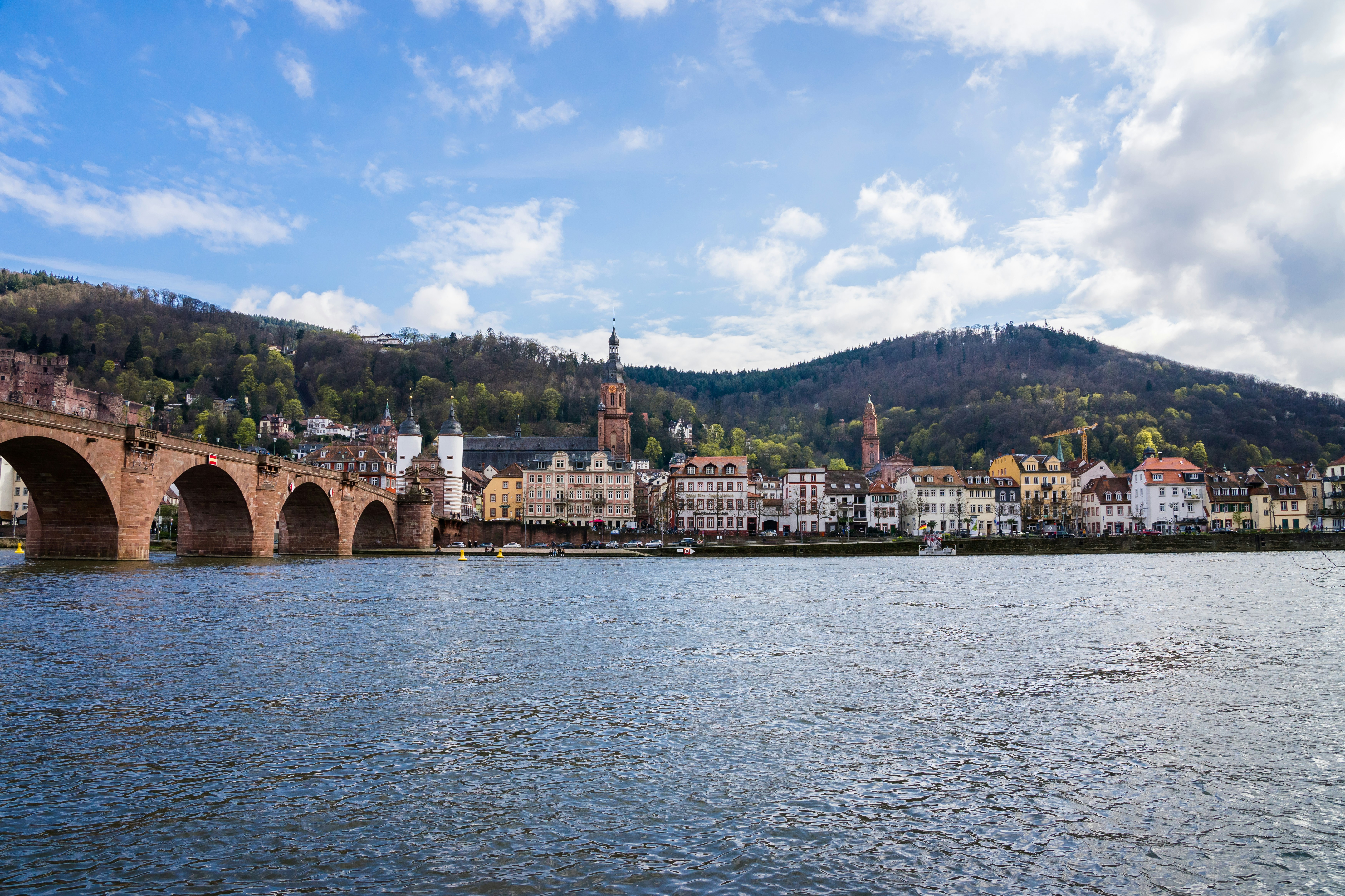 Stone bridge arches gracefully over a wide river with a quaint town and forested hills in the background under a partly cloudy sky.