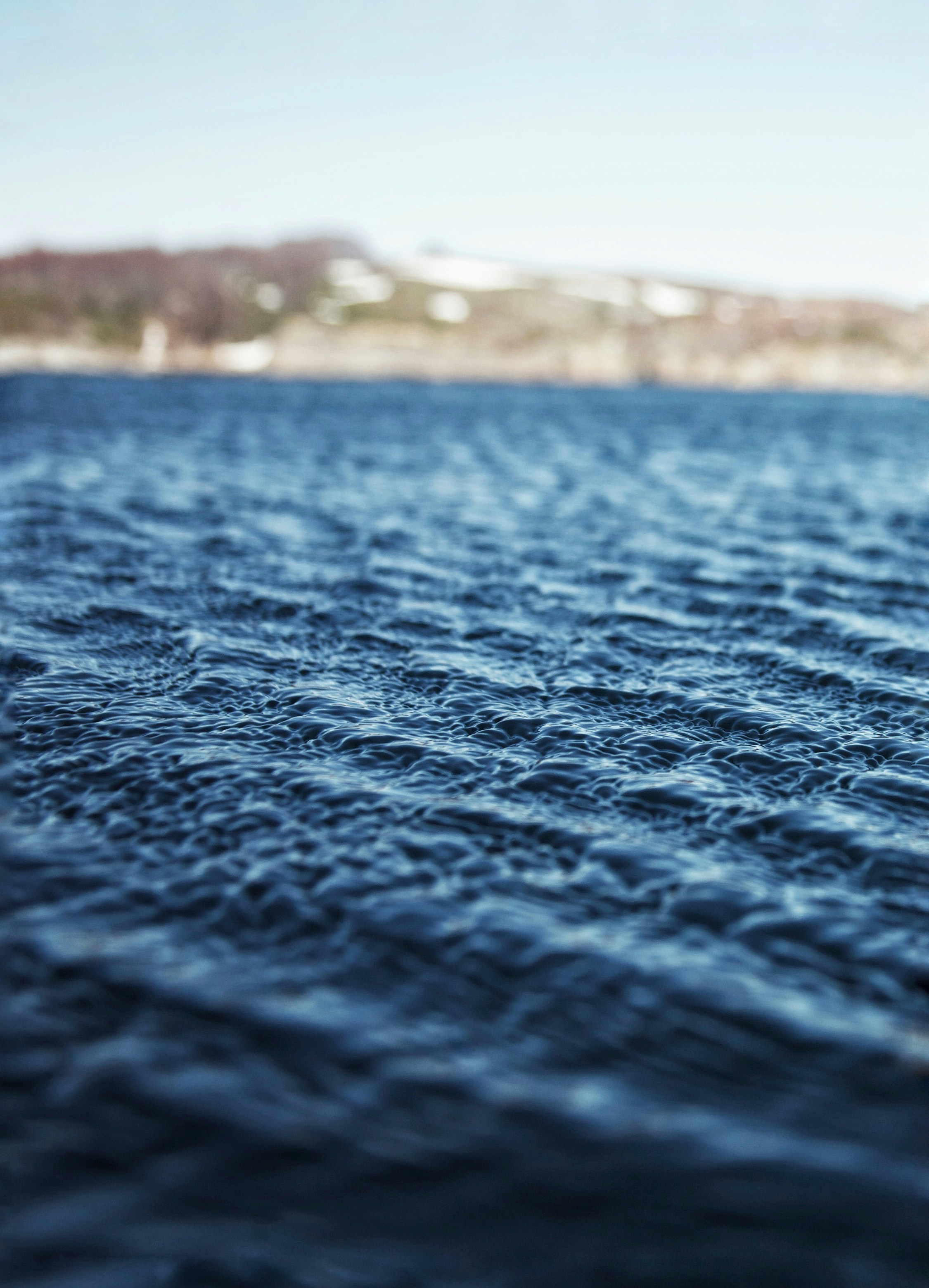 Close-up view of a tranquil water surface, showcasing gentle ripples under a clear sky. The background hints at distant landforms.