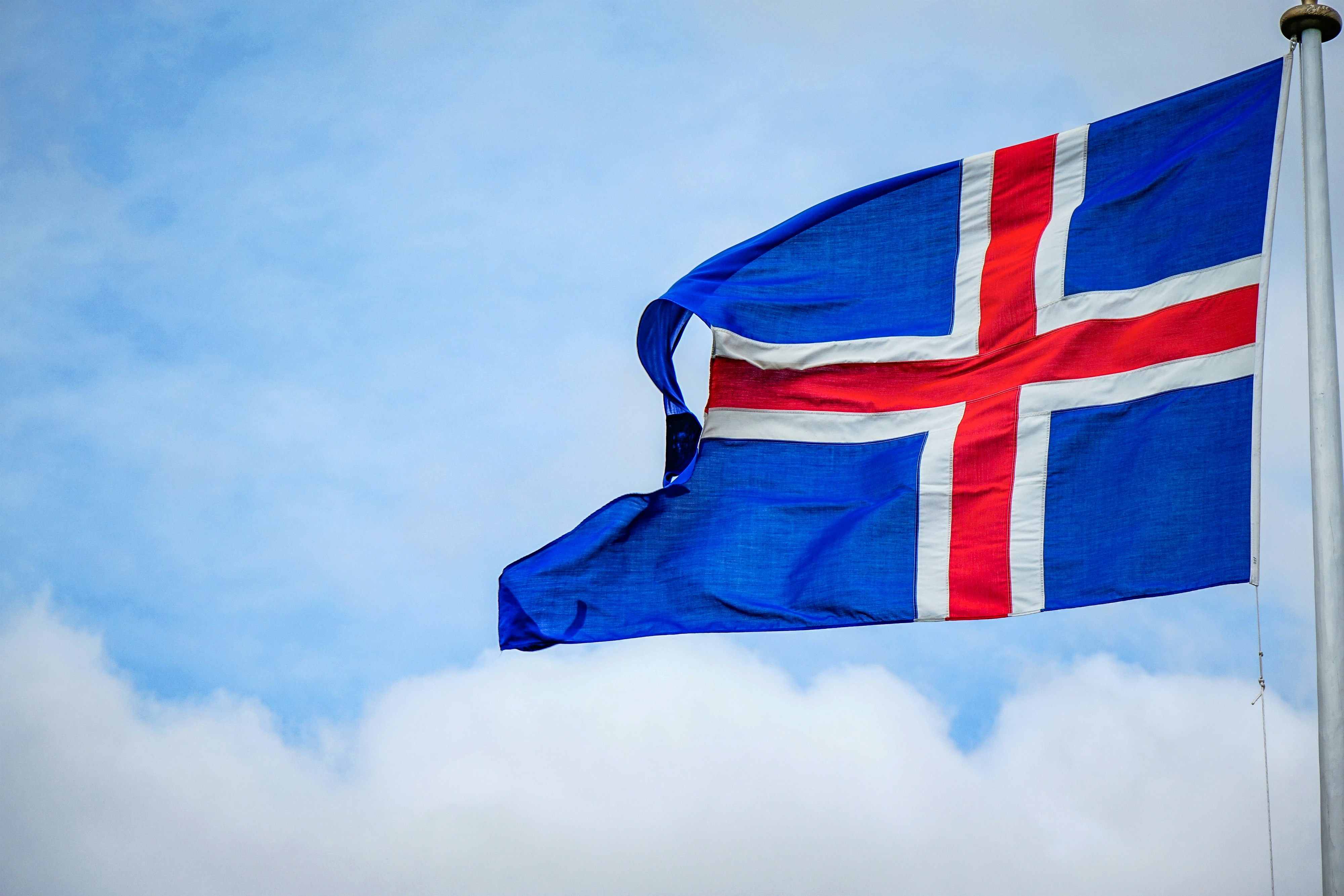 Icelandic flag unfurls on a flagpole against a bright, cloudy blue sky.
