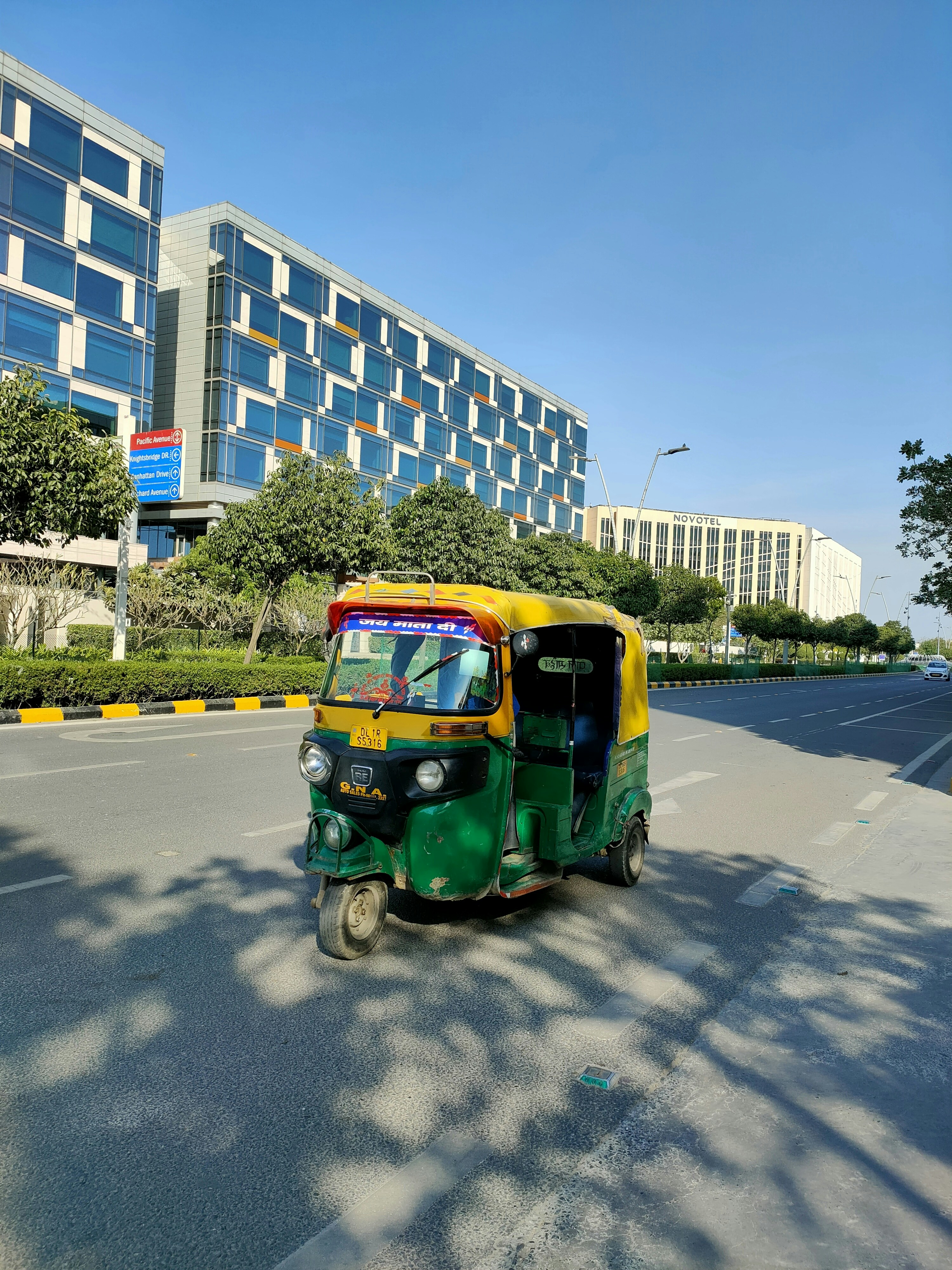 Photograph shows a green-yellow auto-rickshaw cruising a sunlit city avenue, with modern glass-fronted buildings and a clear blue sky.