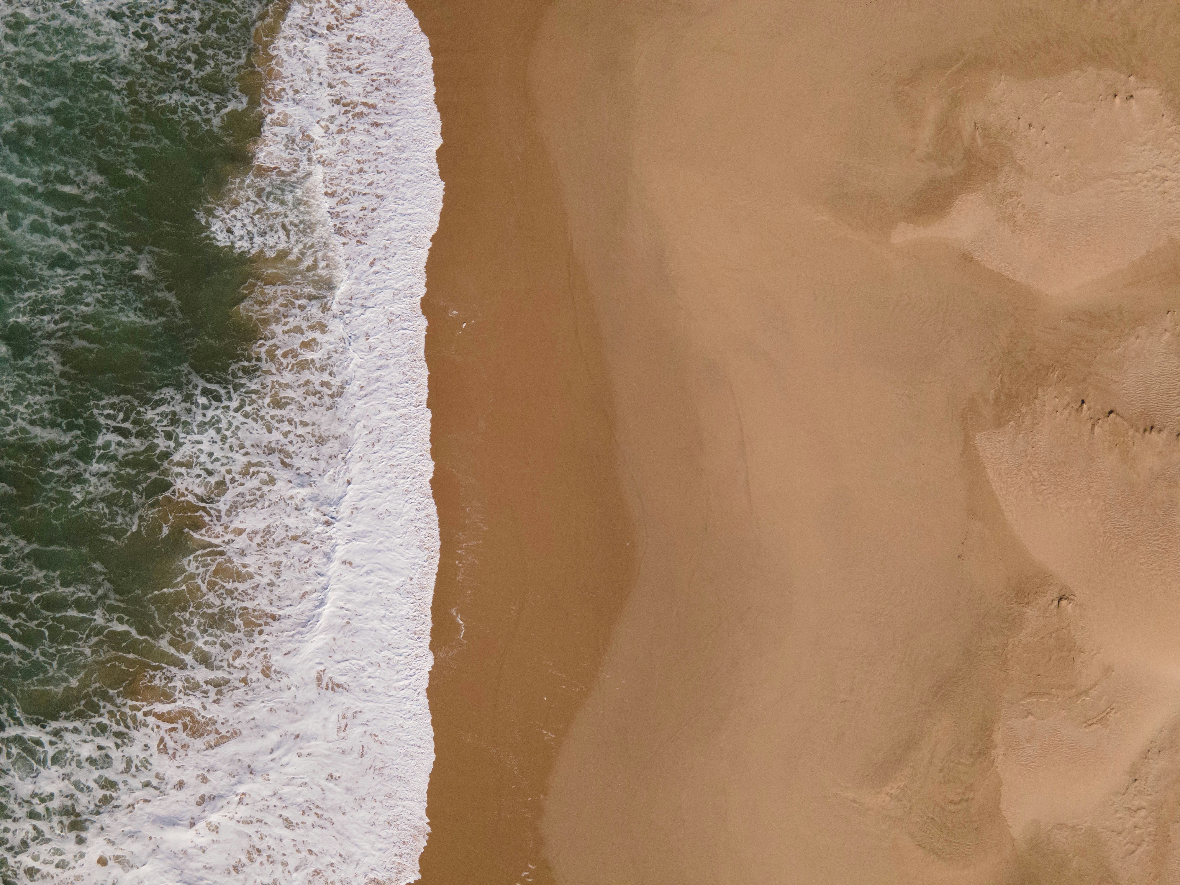 Drone image of green and blue ocean waves with foamy edges meeting a sandy yellow and brown beach.