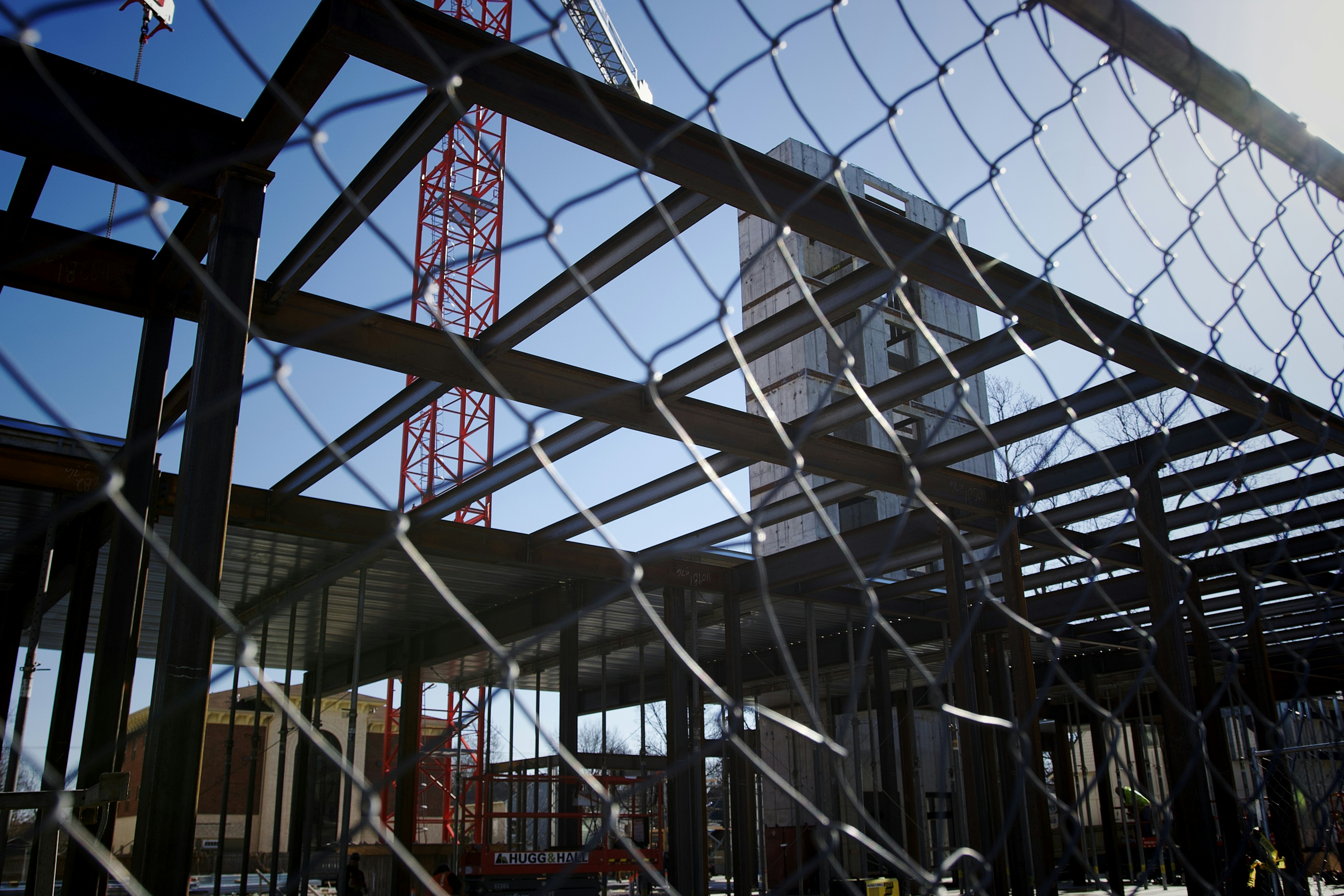 Steel beams and a crane framed by a chain link fence under a clear blue sky.