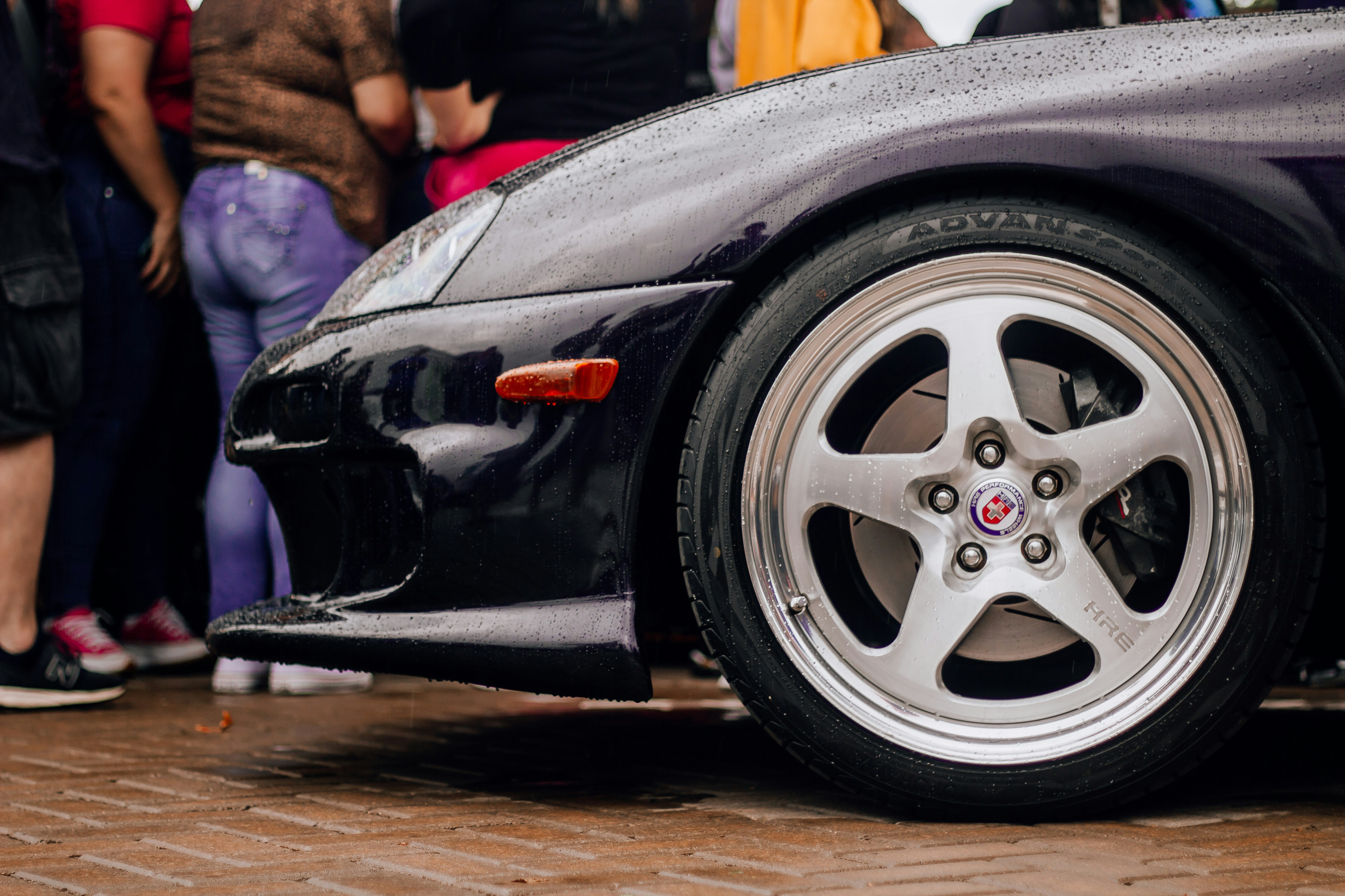 Close-up of a car's front wheel and bumper with a crowd in the background.