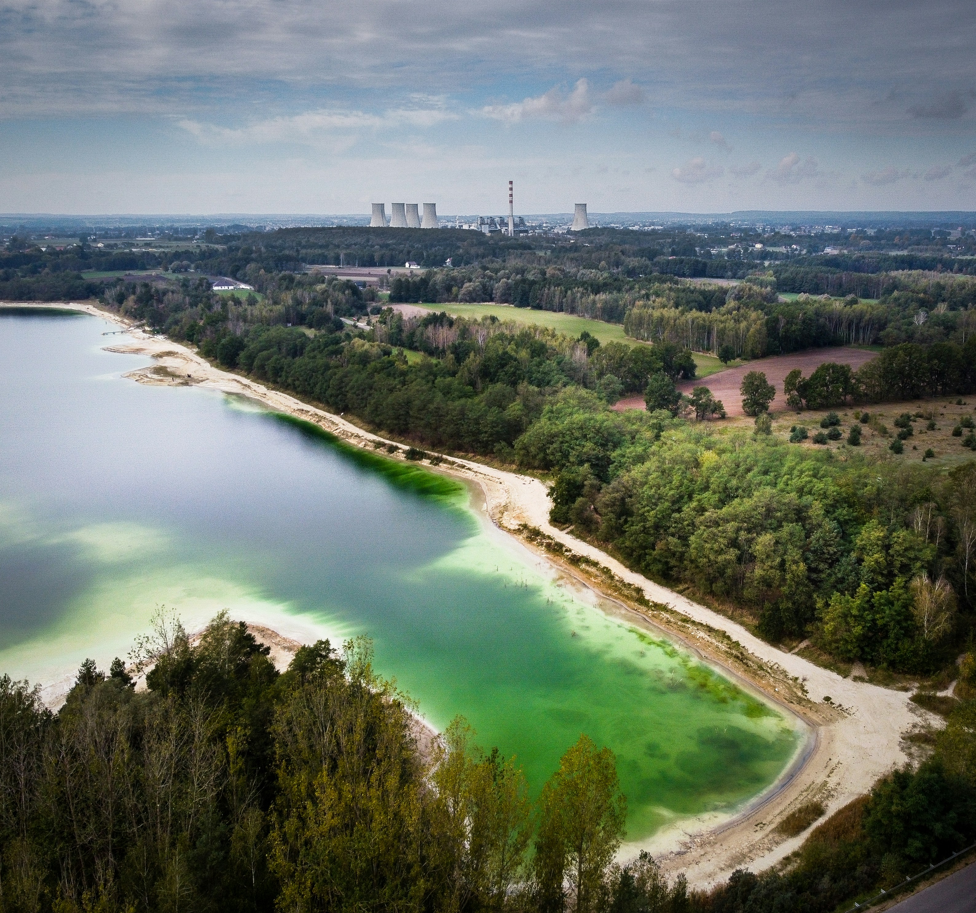 Aerial view of a vibrant green lake surrounded by lush forest with an industrial skyline in the distance.