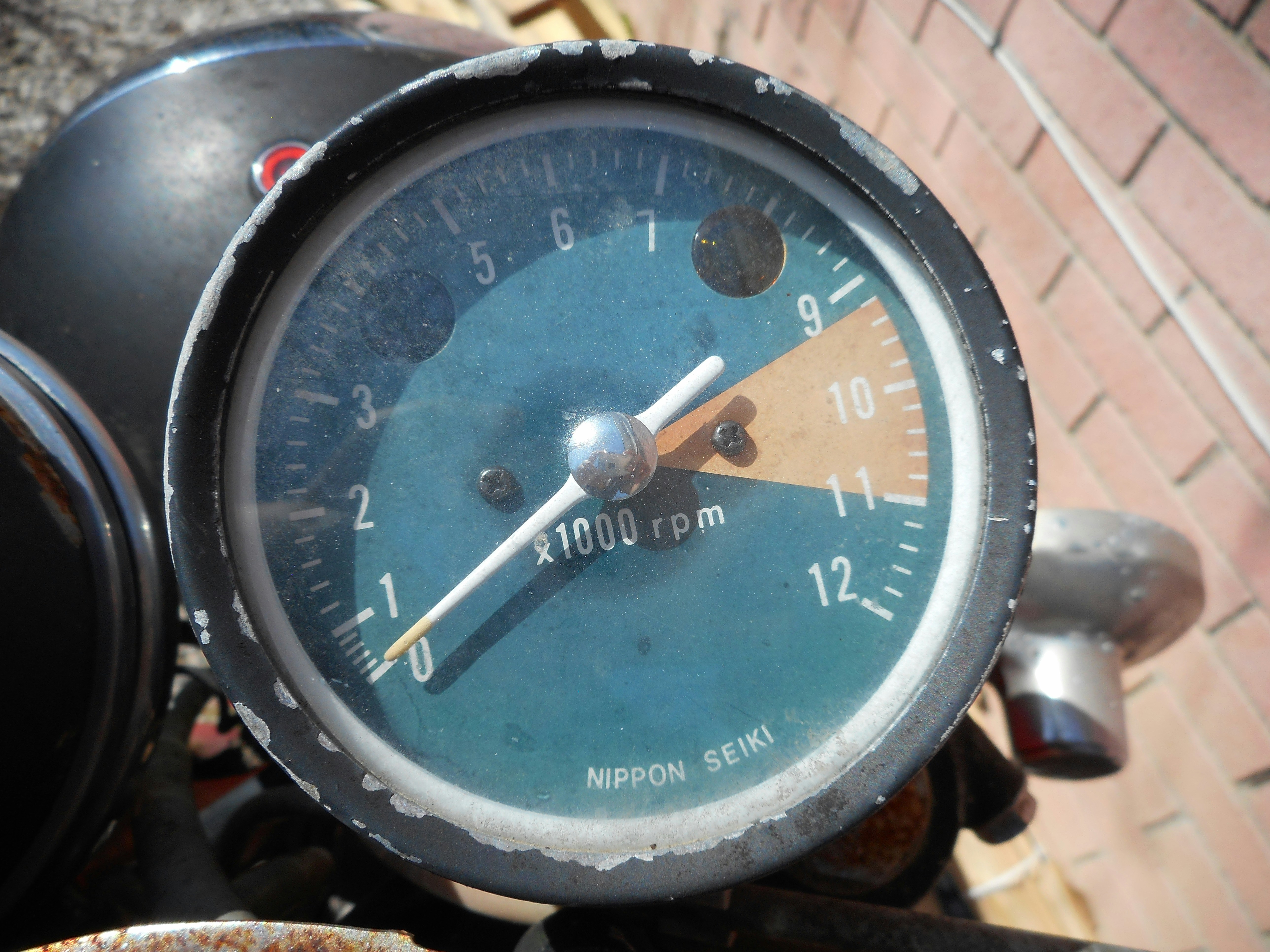 Close-up photograph of a motorcycle tachometer, highlighting the RPM dial with a weathered glass face against a brick-paved background.