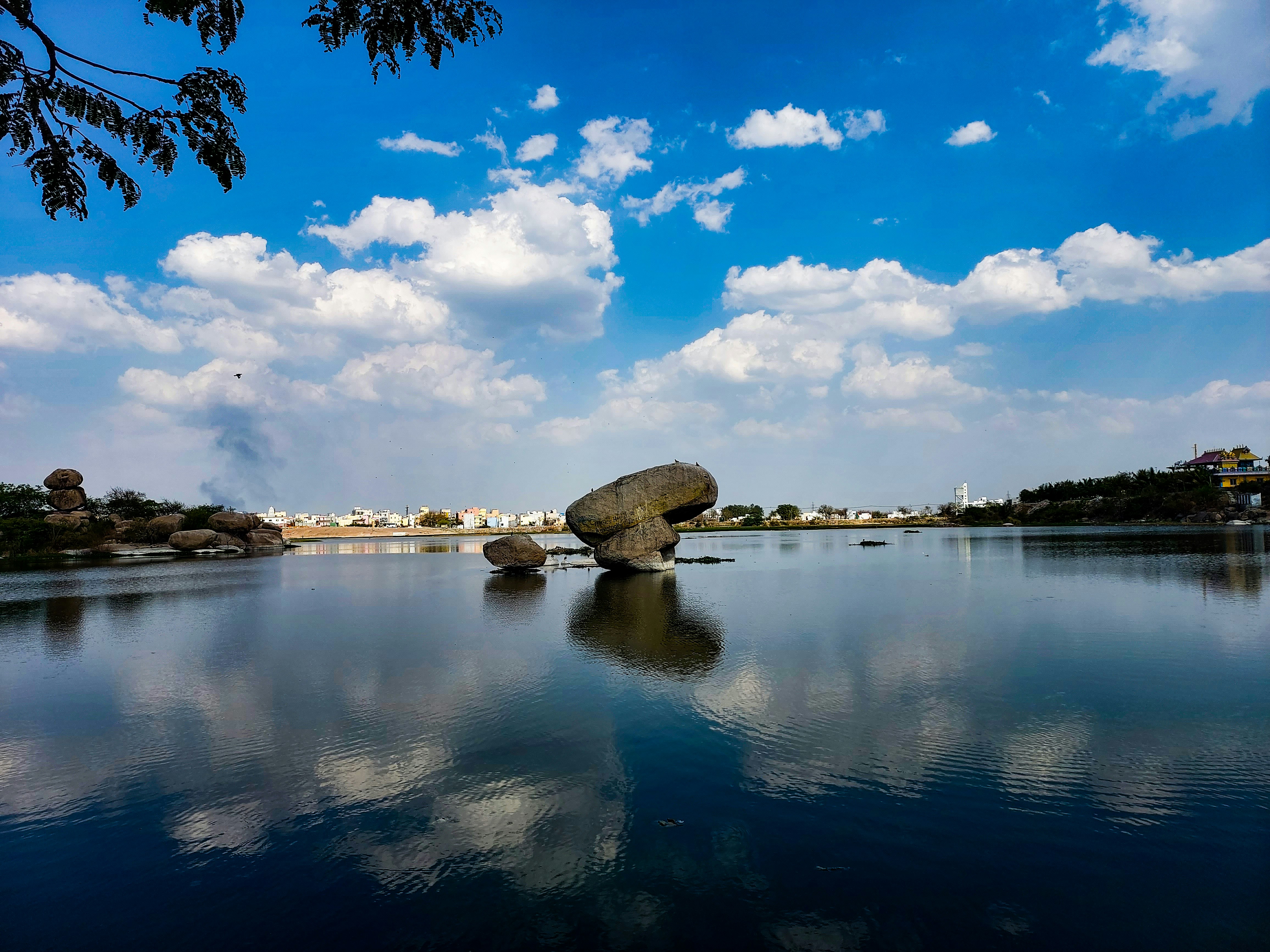 Large rock balancing in the center of a serene lake under a bright blue sky with scattered clouds.