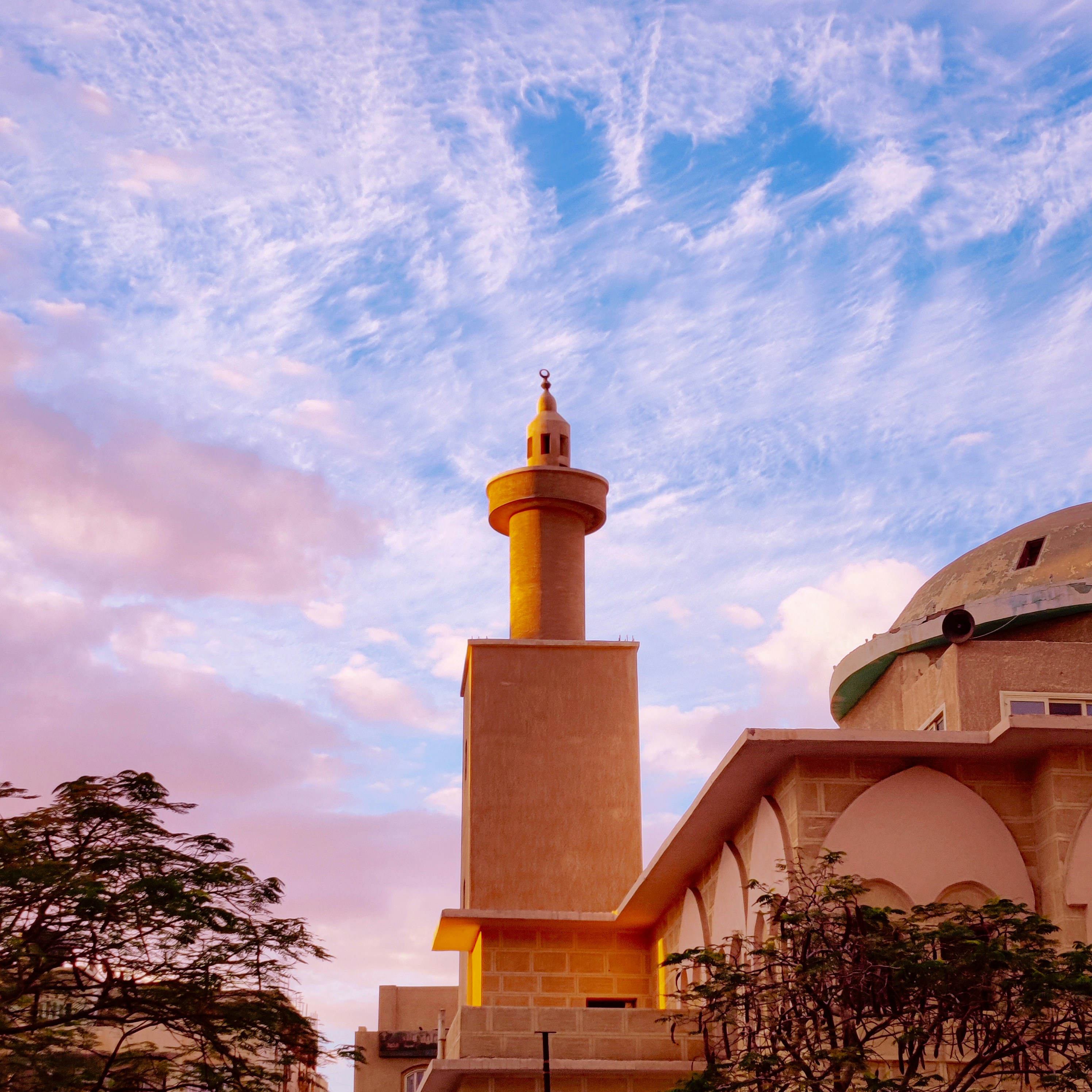 Warm sandstone minaret rises beside a domed building, with trees in the foreground. A pastel sky streaked with pink clouds provides a calm backdrop.