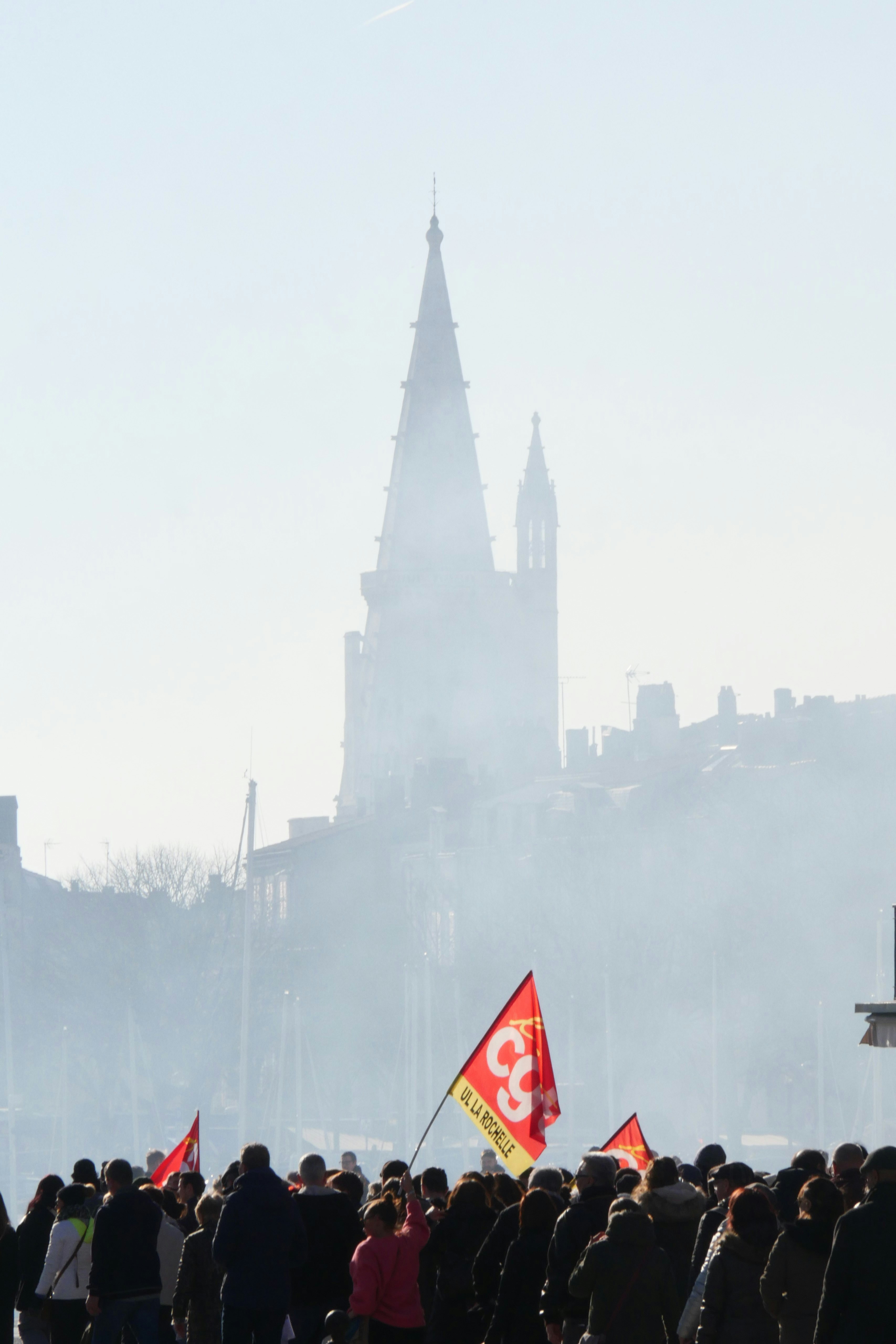 Crowd fills a city square as red flags rise, with a tall Gothic spire fading into morning haze.