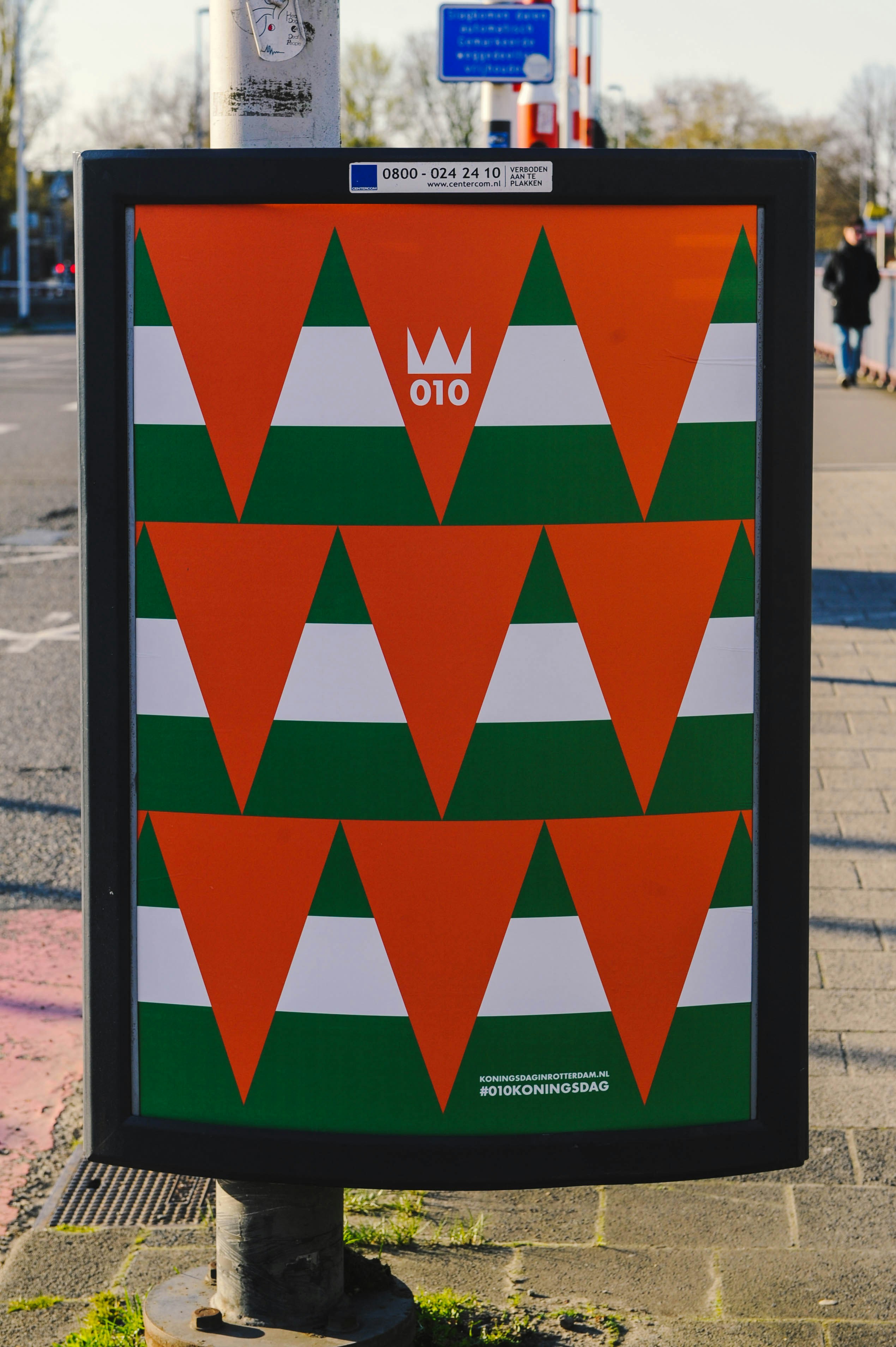 Graphic poster on a street panel featuring bold orange triangles against green shapes, with a crown logo at the top and a campaign hashtag at the bottom.