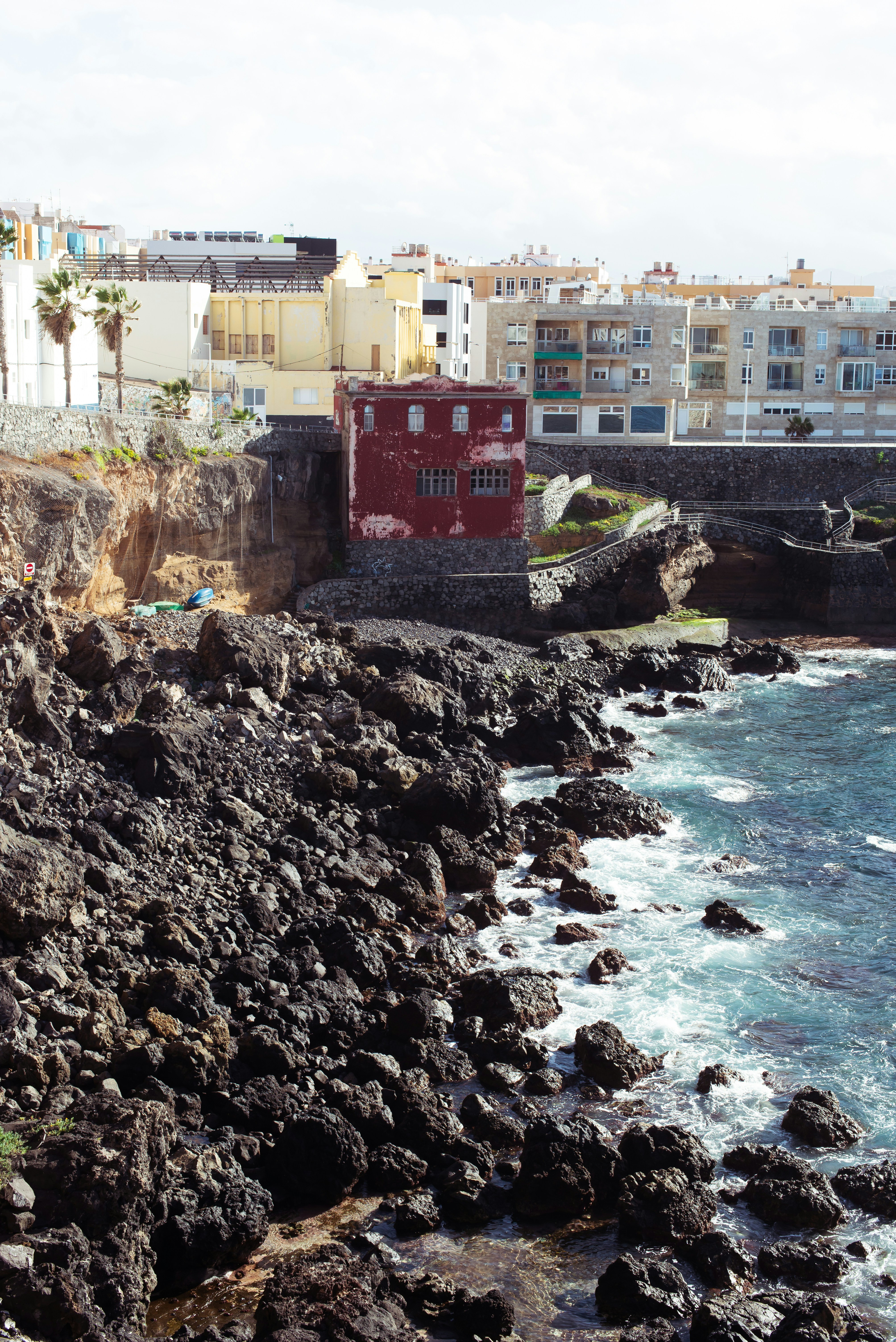 Weathered red building perched on rocky coastline, surrounded by crashing waves and urban backdrop. The scene captures a blend of nature and human history.