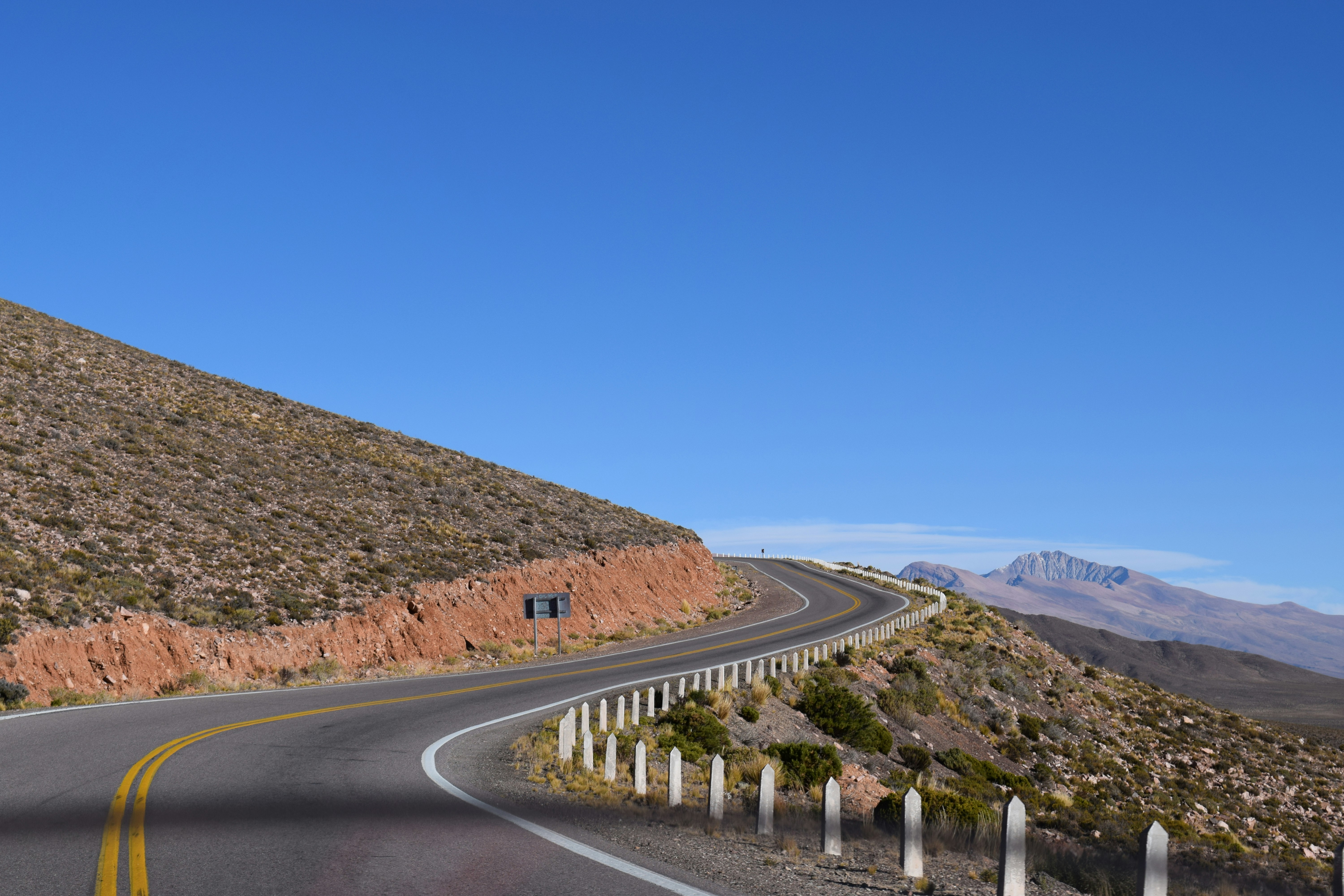 Curving road meanders through arid terrain under a clear blue sky, leading toward distant mountains.