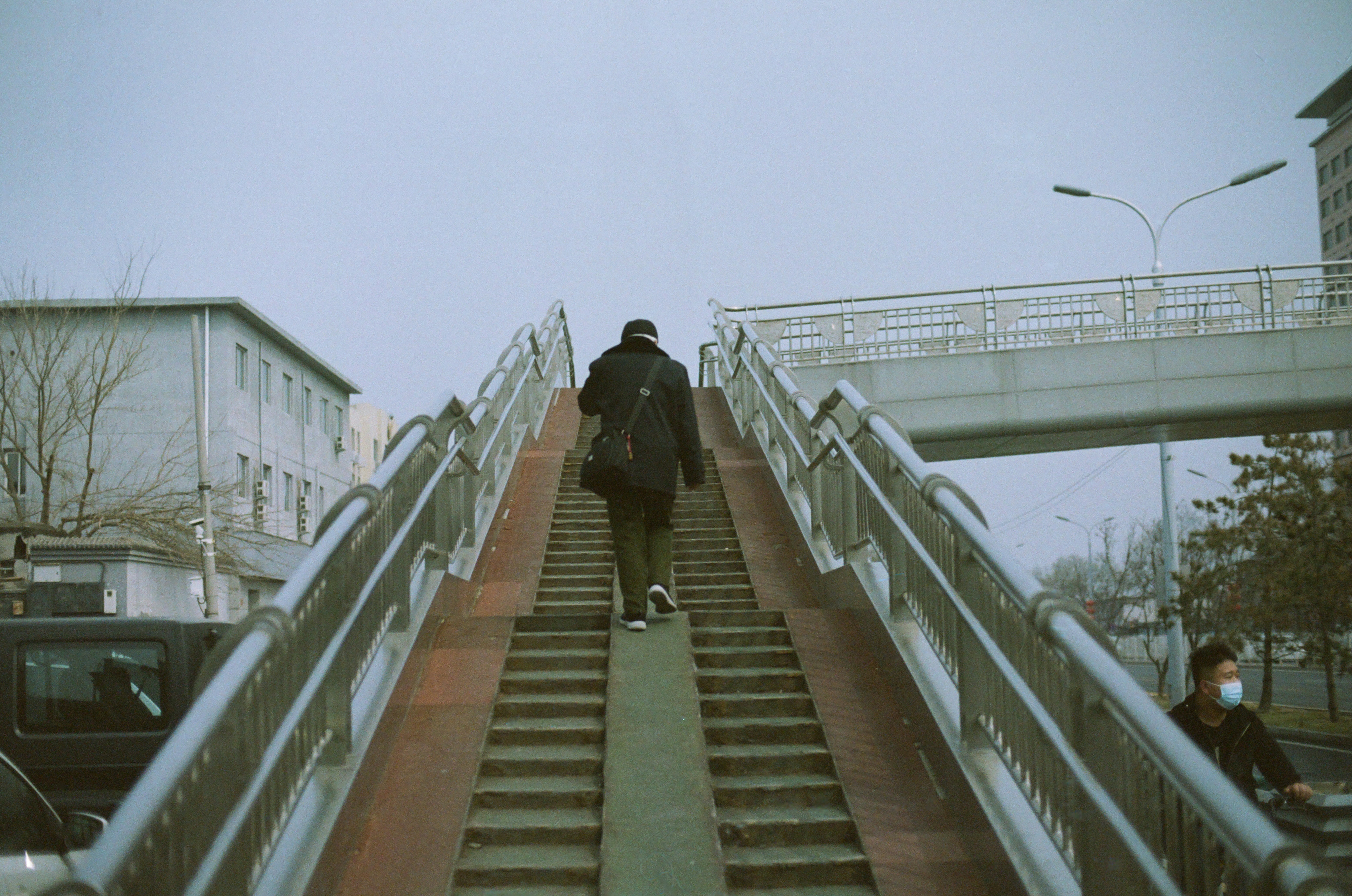 A solitary figure ascends an urban overpass under a gray sky, juxtaposed with a bustling street below.