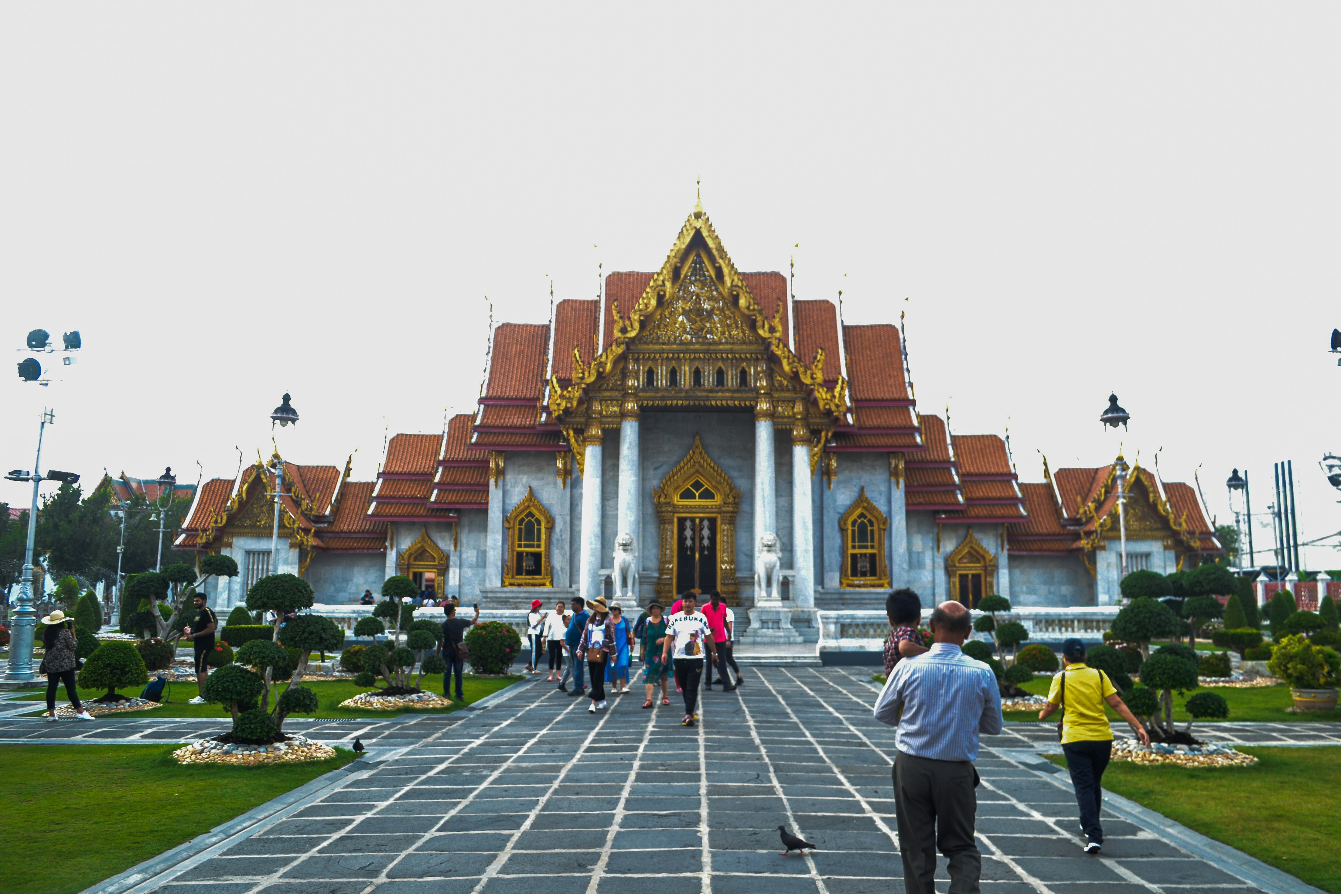Visitors strolling towards an ornate temple with intricate gold details and manicured gardens. The scene captures a vibrant cultural moment.