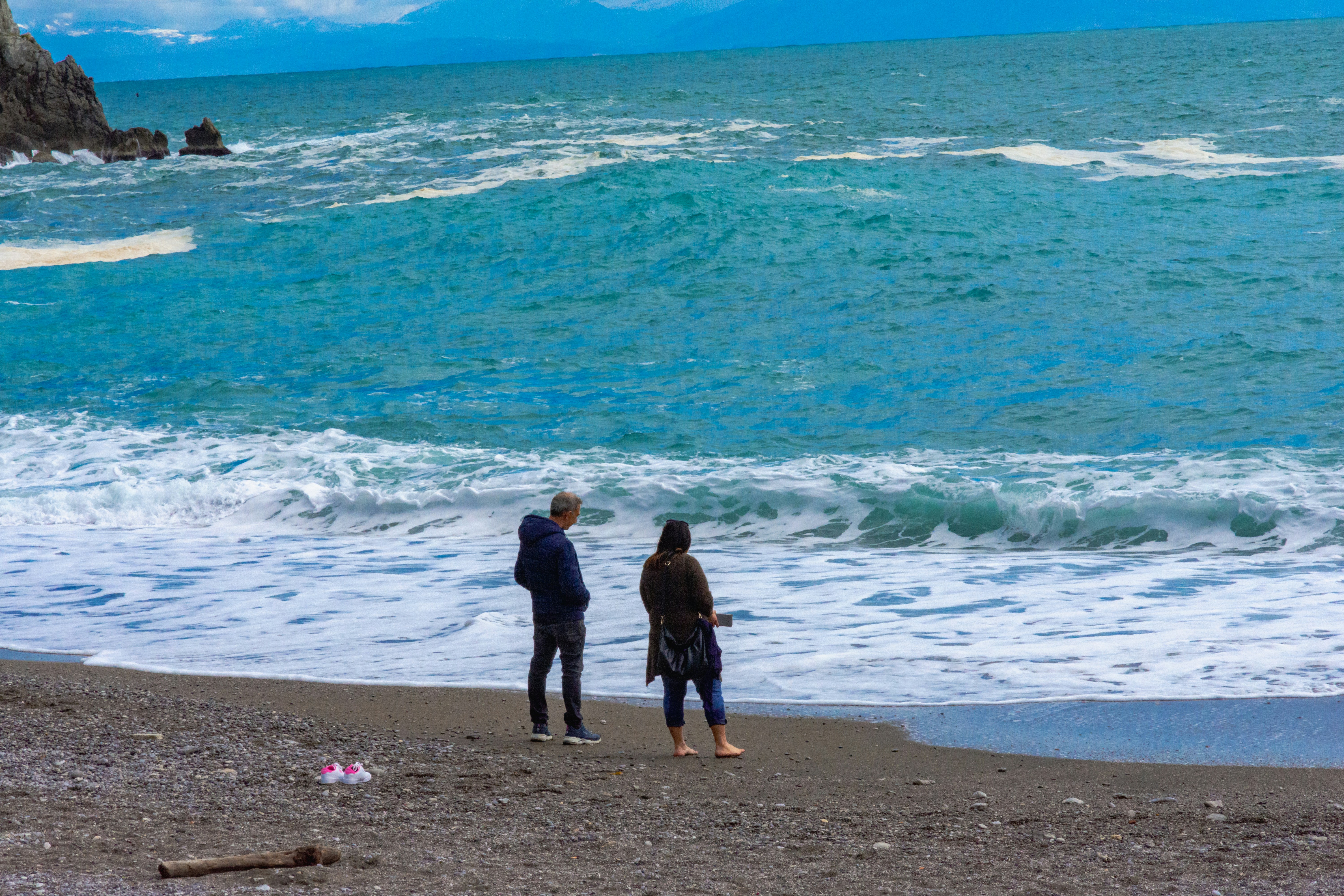 Two people stand on a pebbled beach gazing at the turquoise ocean under a cloudy sky.