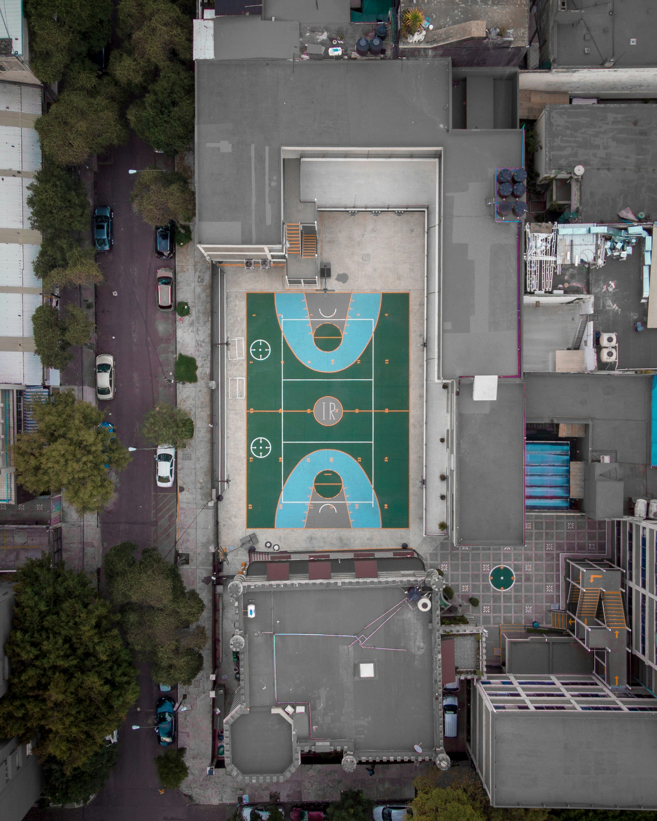 Aerial Portrait of a Neon-Turquoise Rooftop Basketball Court Amid Dense Urban Rooftops