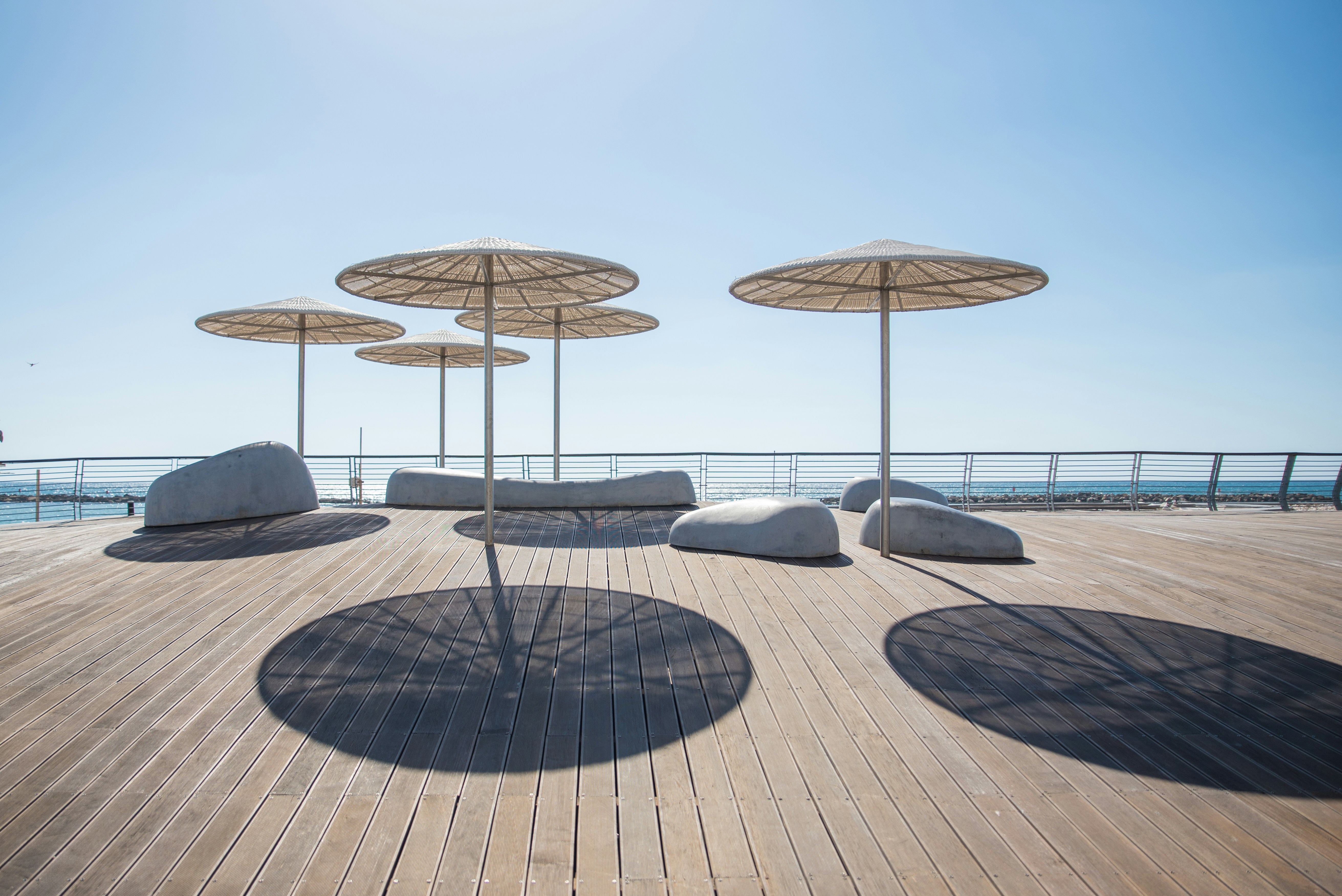 Photograph of a sunlit seaside boardwalk with circular parasols casting large shadows on the wooden planks, a railing and calm sea on the horizon.