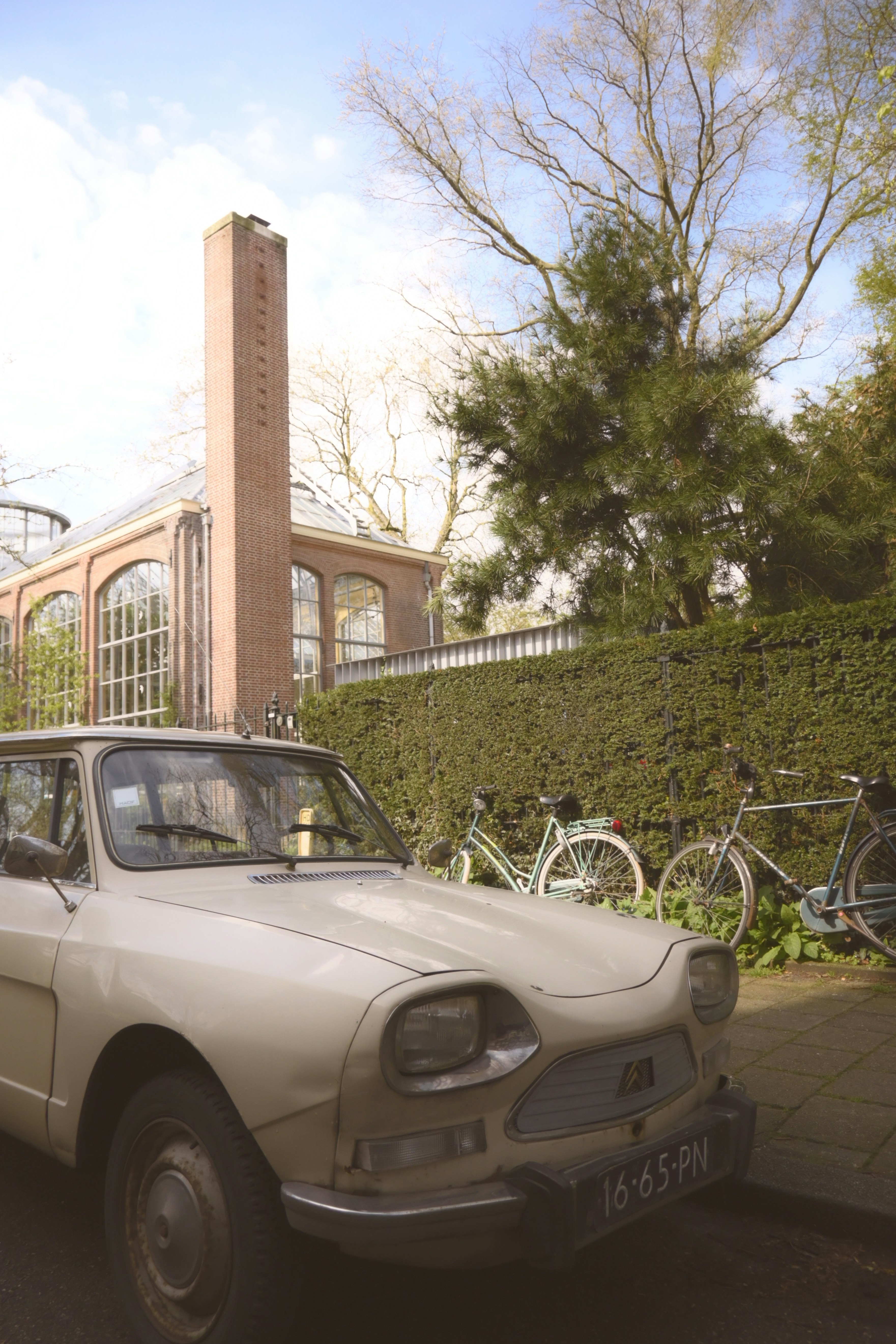 Vintage Renault on a Sunlit Street: Bicycles, Hedge, and Brick Conservatory