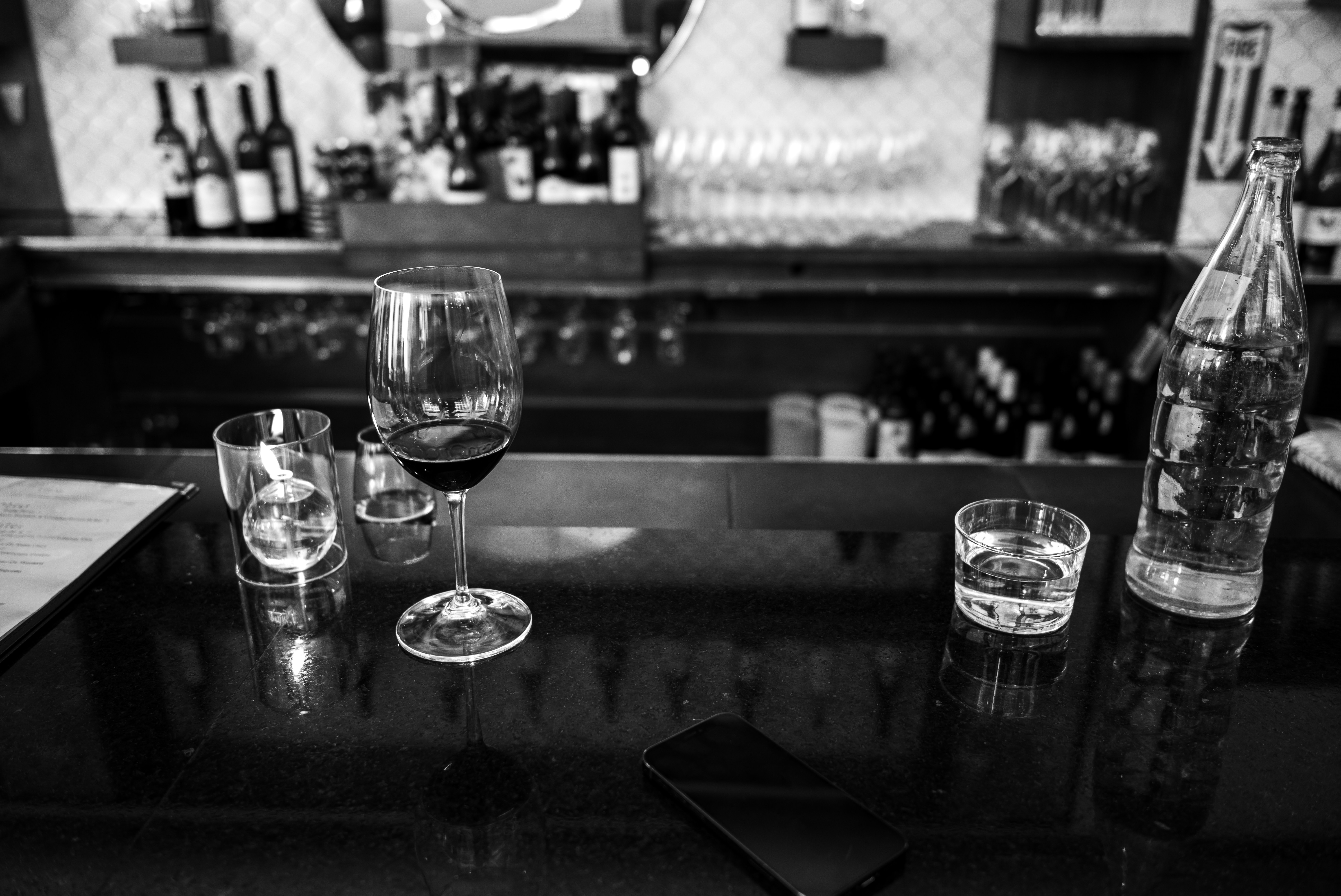 Black and white bar scene with empty glasses and a wine bottle on a reflective counter.