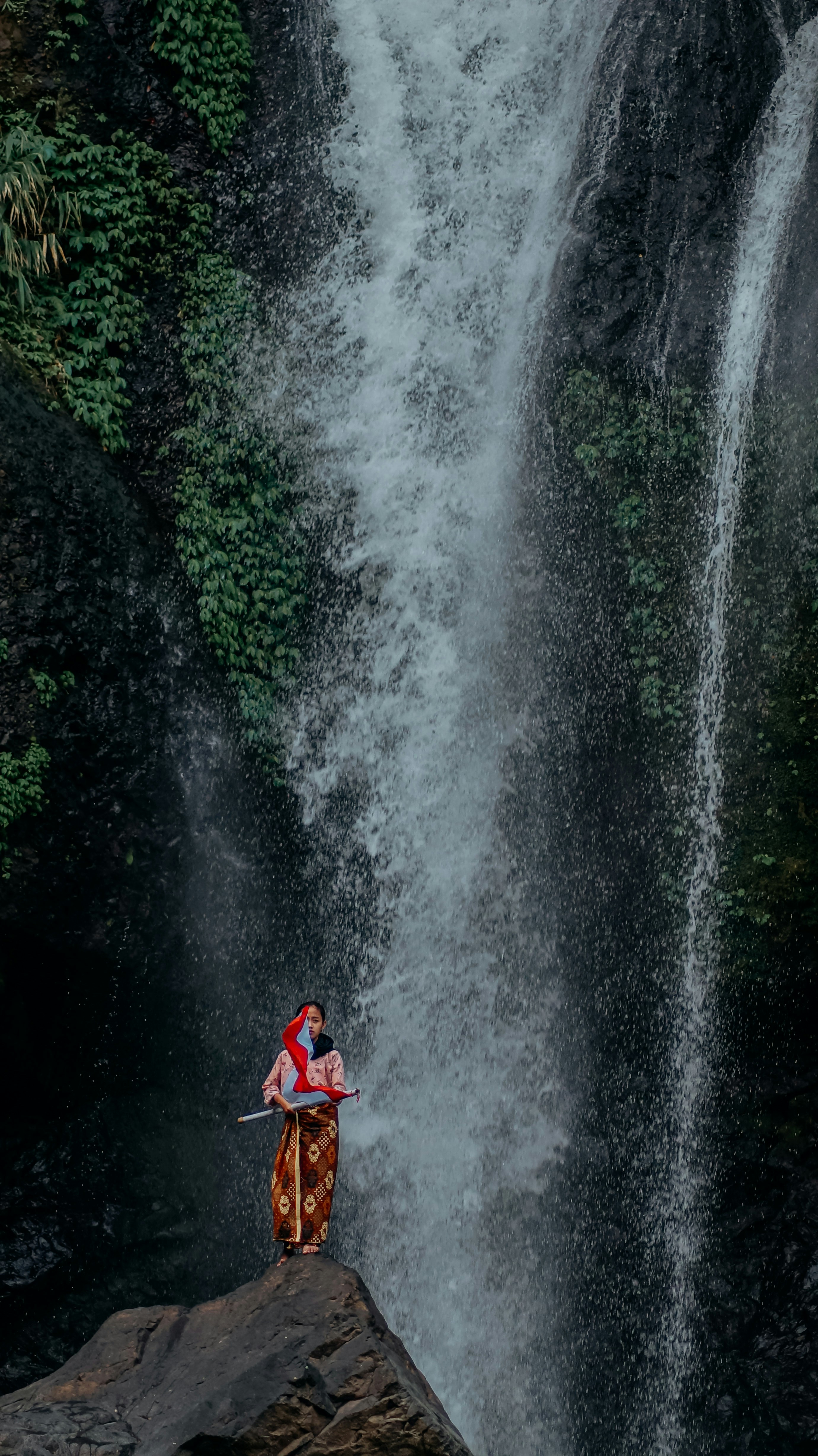 A lone performer in batik attire stands on a rock at the base of a tall waterfall, framed by lush greenery and mist.