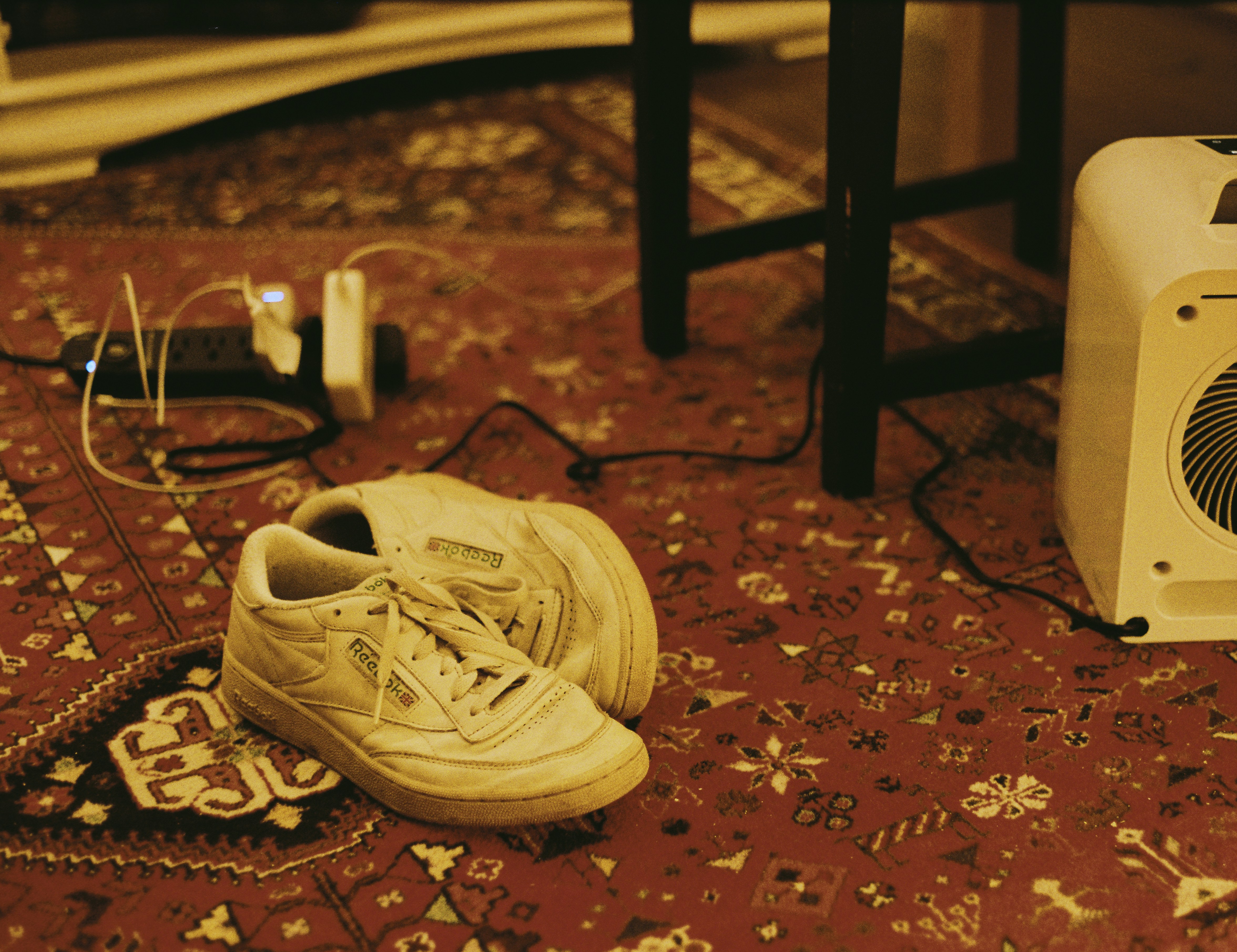 Worn white sneakers rest on a patterned rug beside a small stereo and loose cords in a warm, amber-lit room.