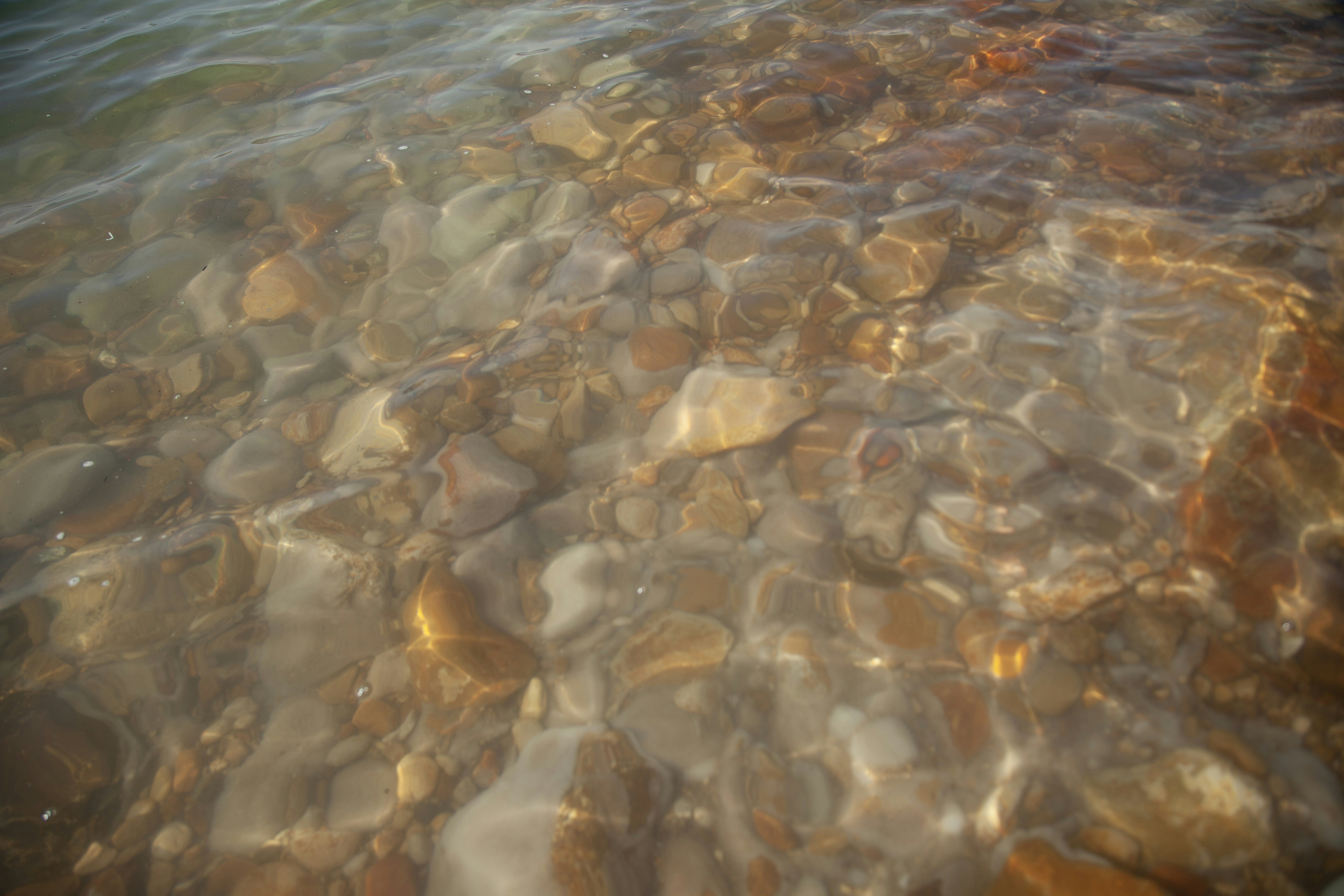 Clear water revealing a mosaic of smooth pebbles beneath the surface, illuminated by gentle sunlight. The tranquil scene invites reflection.