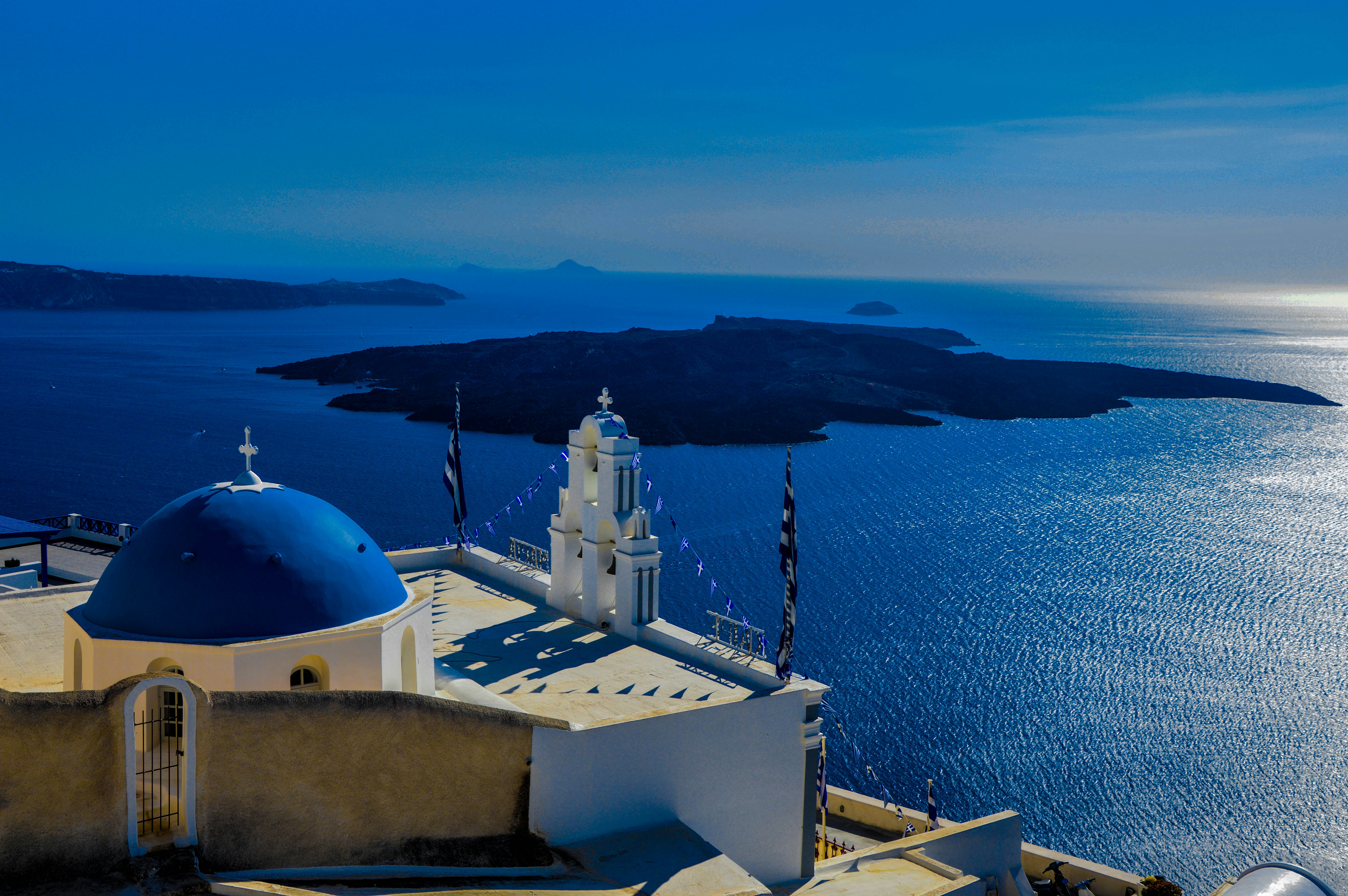 Blue-domed church overlooking the shimmering Aegean Sea under a clear sky.