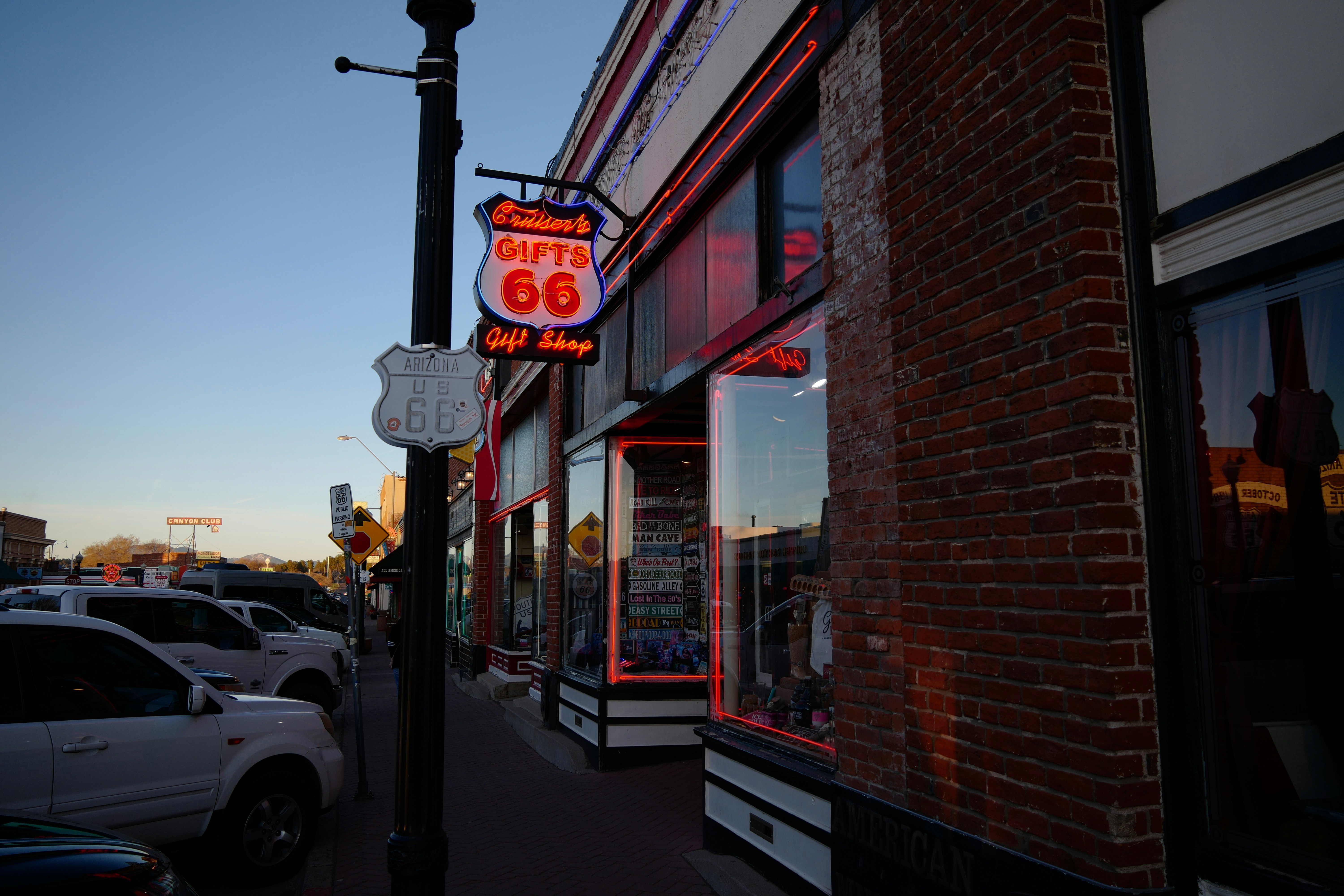 Neon Route 66 sign on a brick storefront glows beside reflective windows as cars line a small-town street at dusk.
